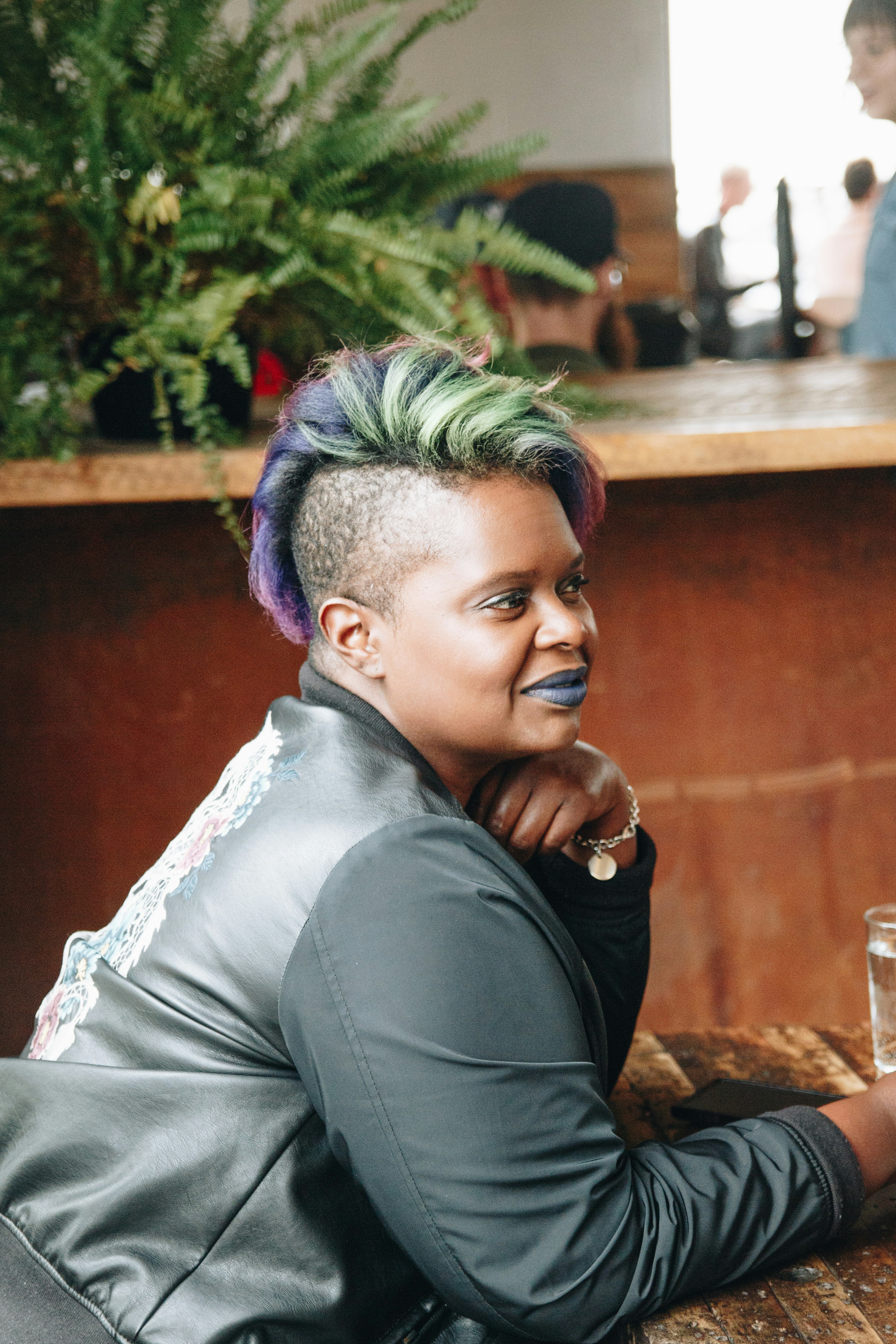 Individual with colorful hair and bold makeup lounging at a rustic table in a lively café atmosphere.