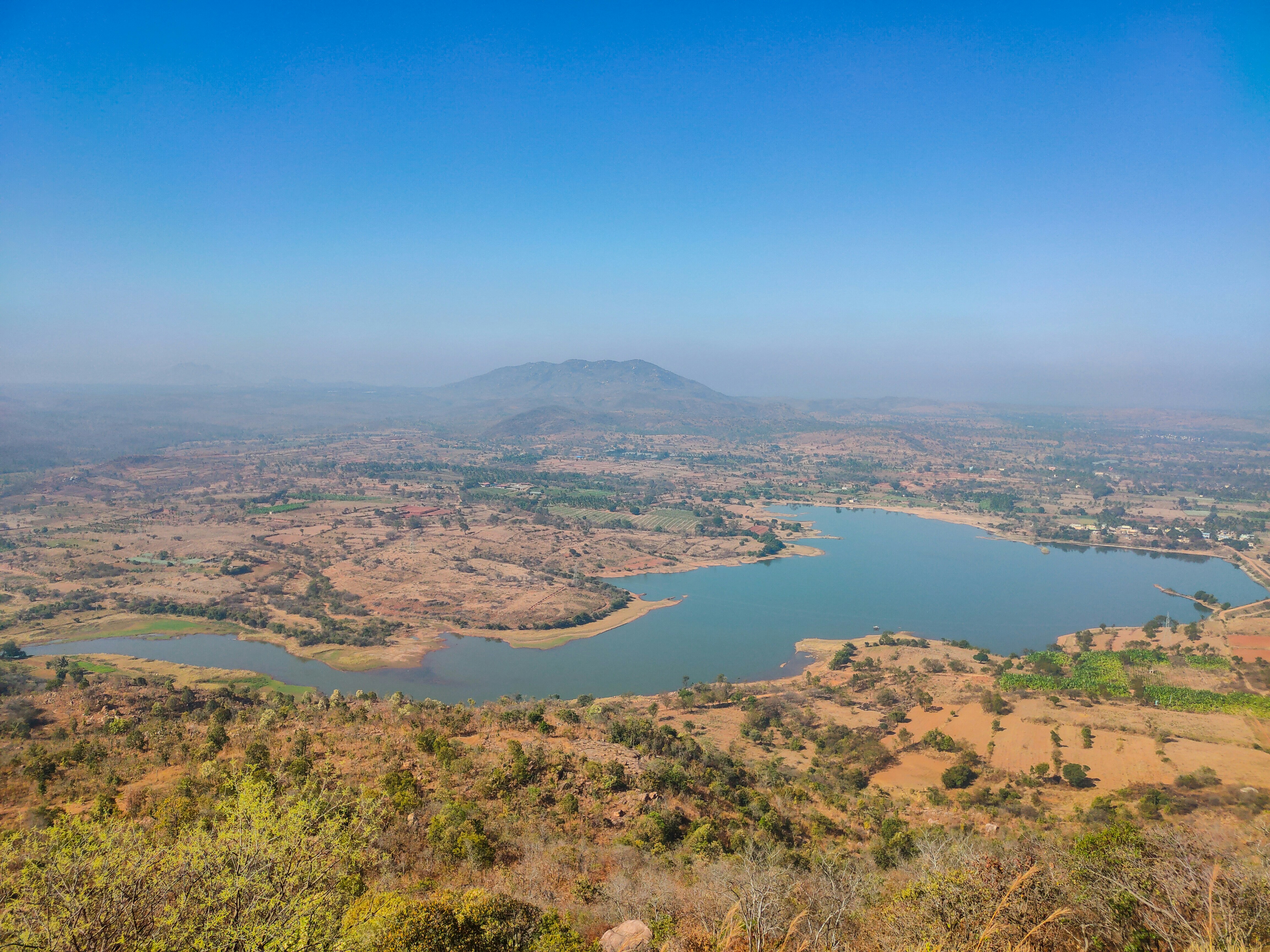 Photograph of a vast landscape featuring a winding lake across a dry valley, with a distant mountain rising on the horizon. A crisp, blue sky completes the tranquil panorama.