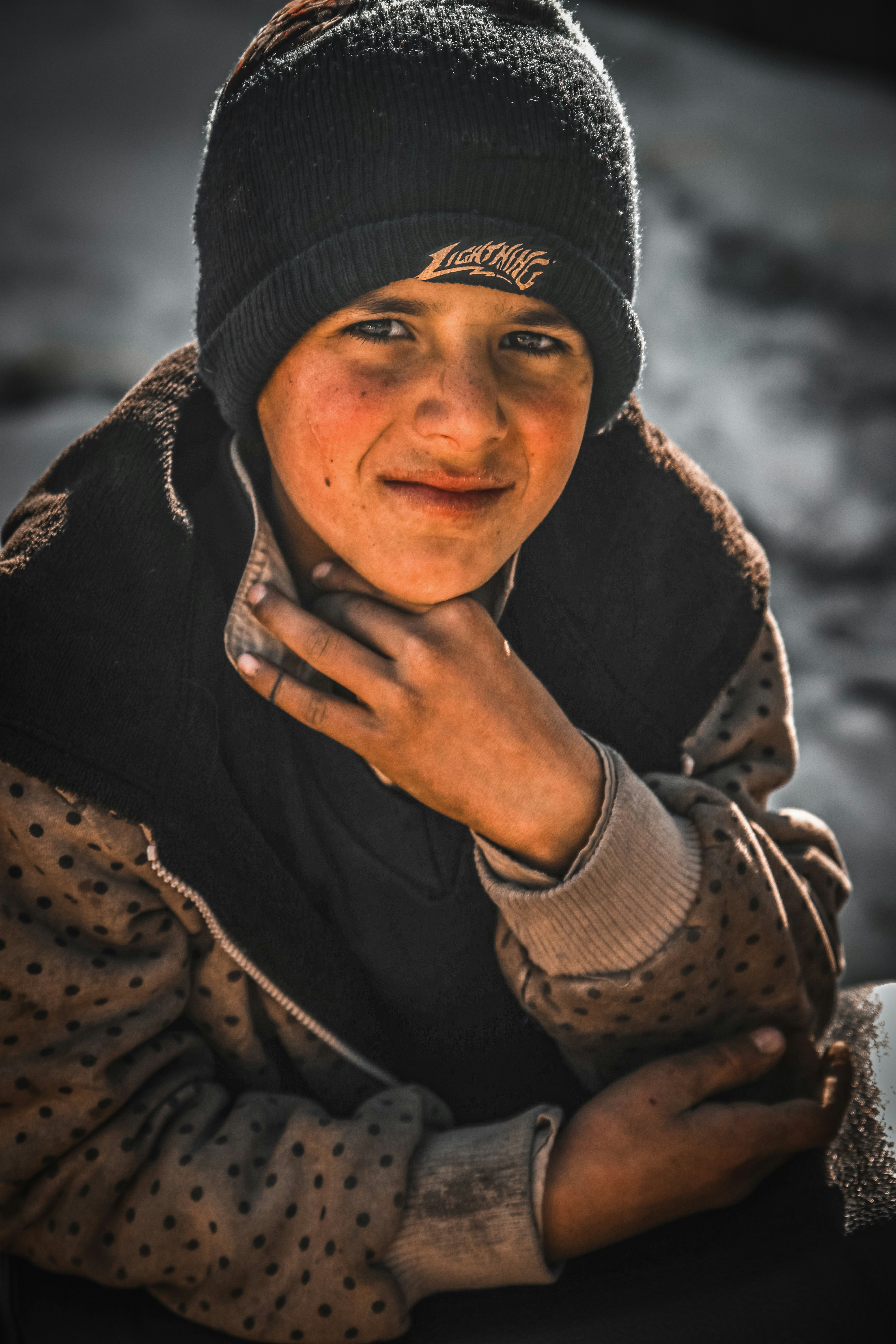 A young boy in a knitted beanie gazes thoughtfully, his hand resting on his chin, surrounded by a softly blurred background that hints at his environment.