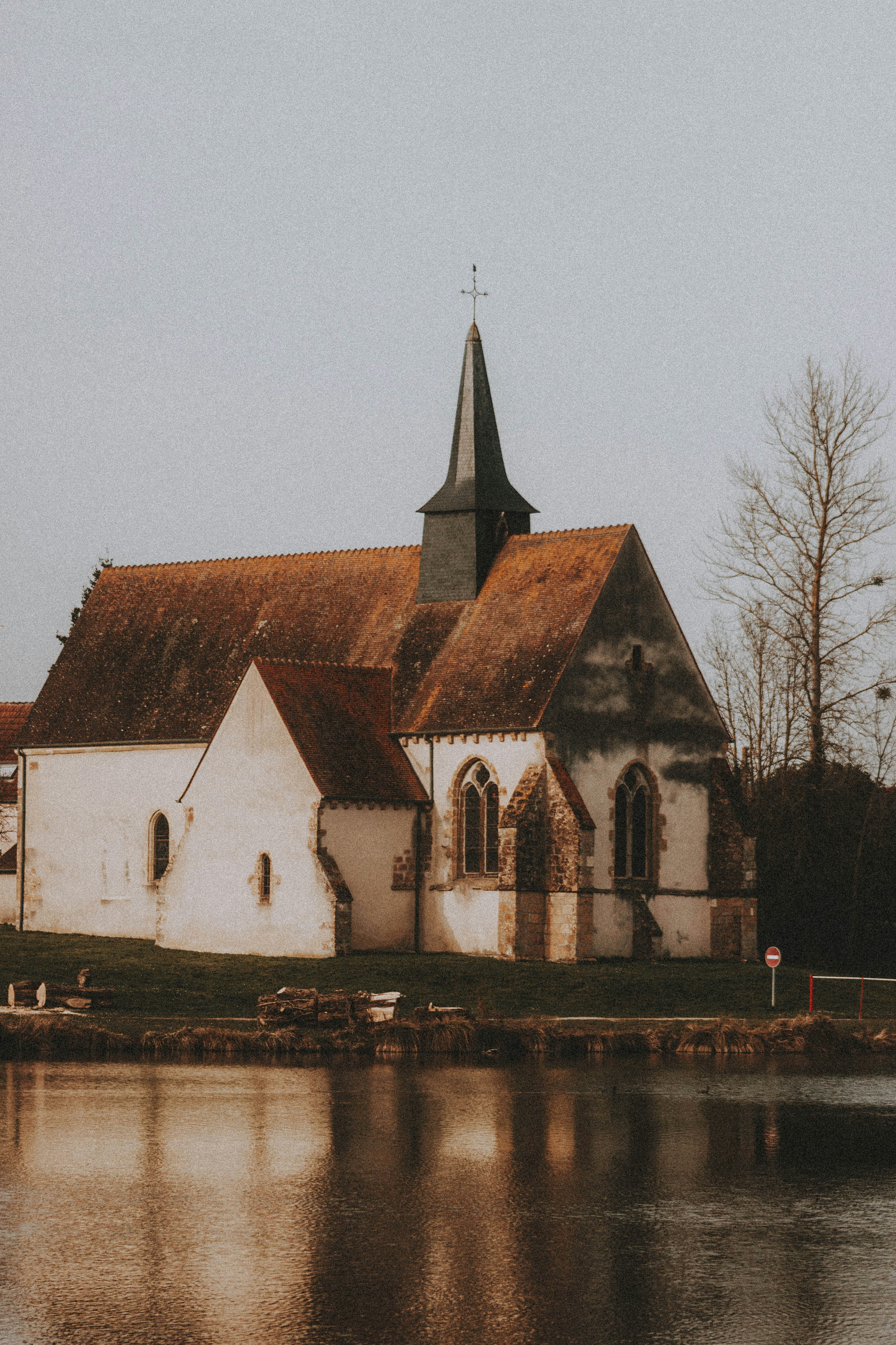 Historic church with a pointed steeple reflected in calm waters, surrounded by bare trees during twilight.