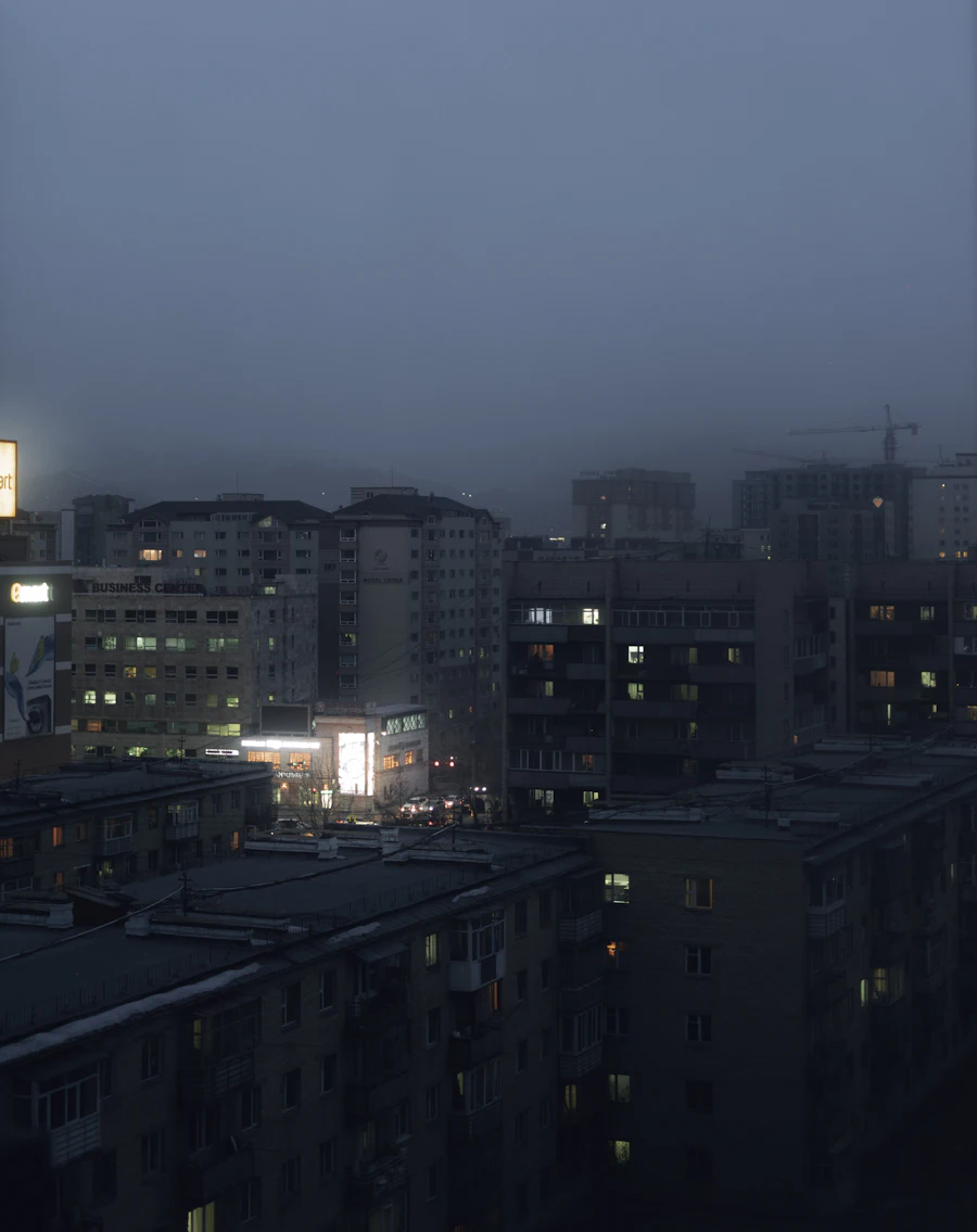 Blue-hour cityscape with dense apartment blocks, glowing windows, and a bright storefront near the center. Fog softens the distant skyline and cranes under an overcast sky.