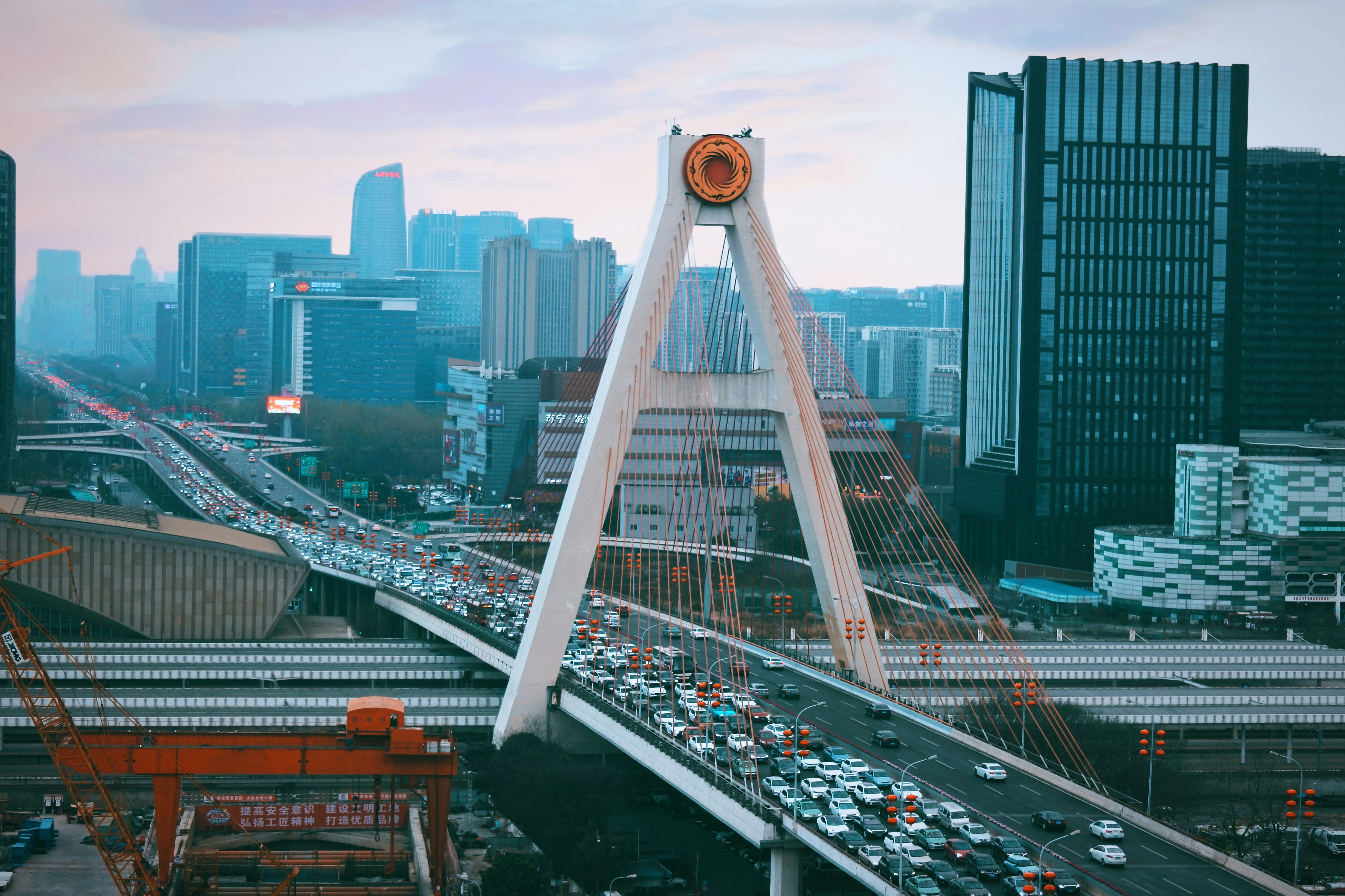 A busy urban overpass featuring a striking cable-stayed bridge amidst a bustling cityscape. The scene captures the rhythm of traffic and architecture at twilight.