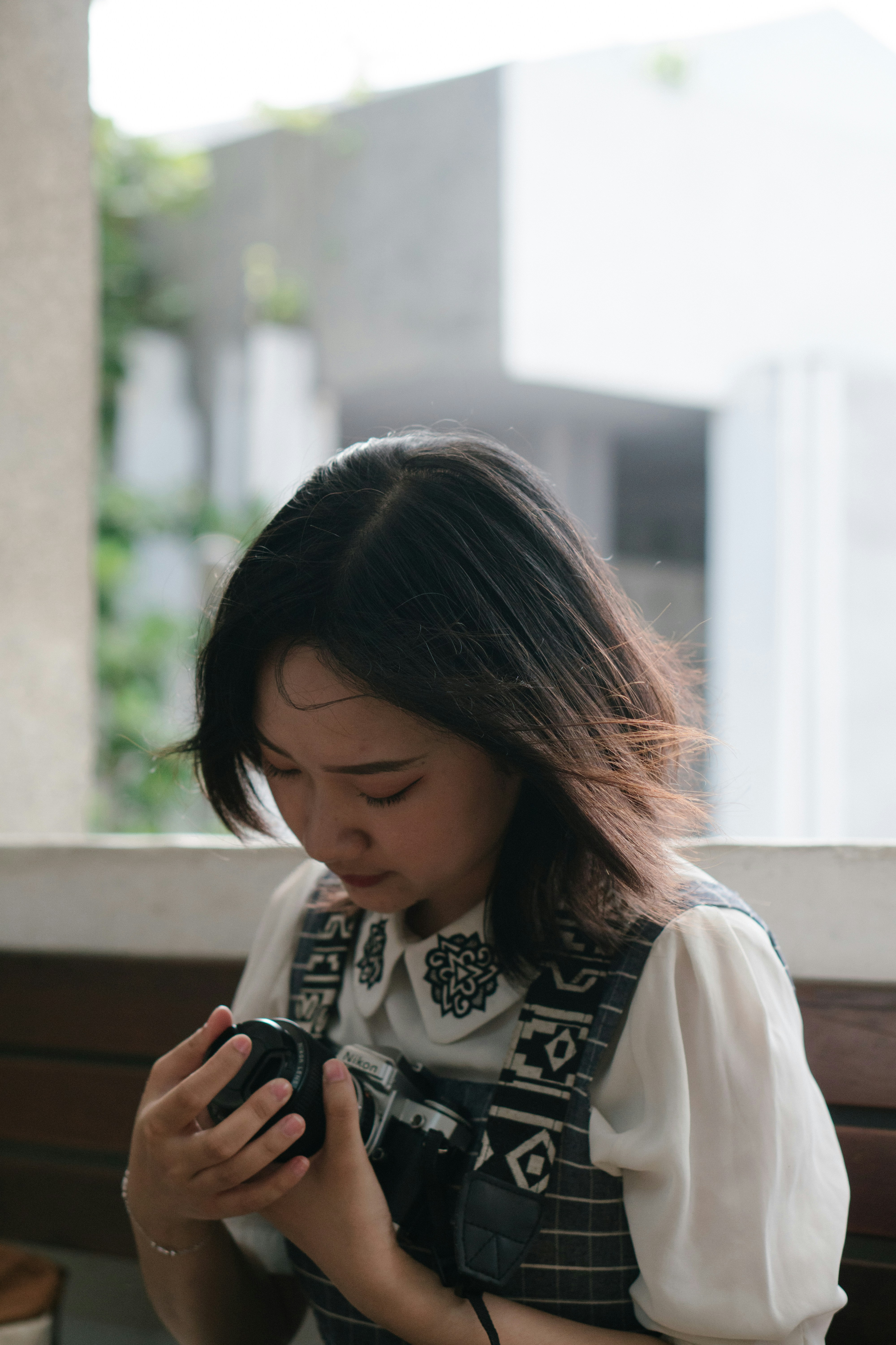 A young woman examines her camera with focused intent, surrounded by soft natural light and minimalistic architecture.