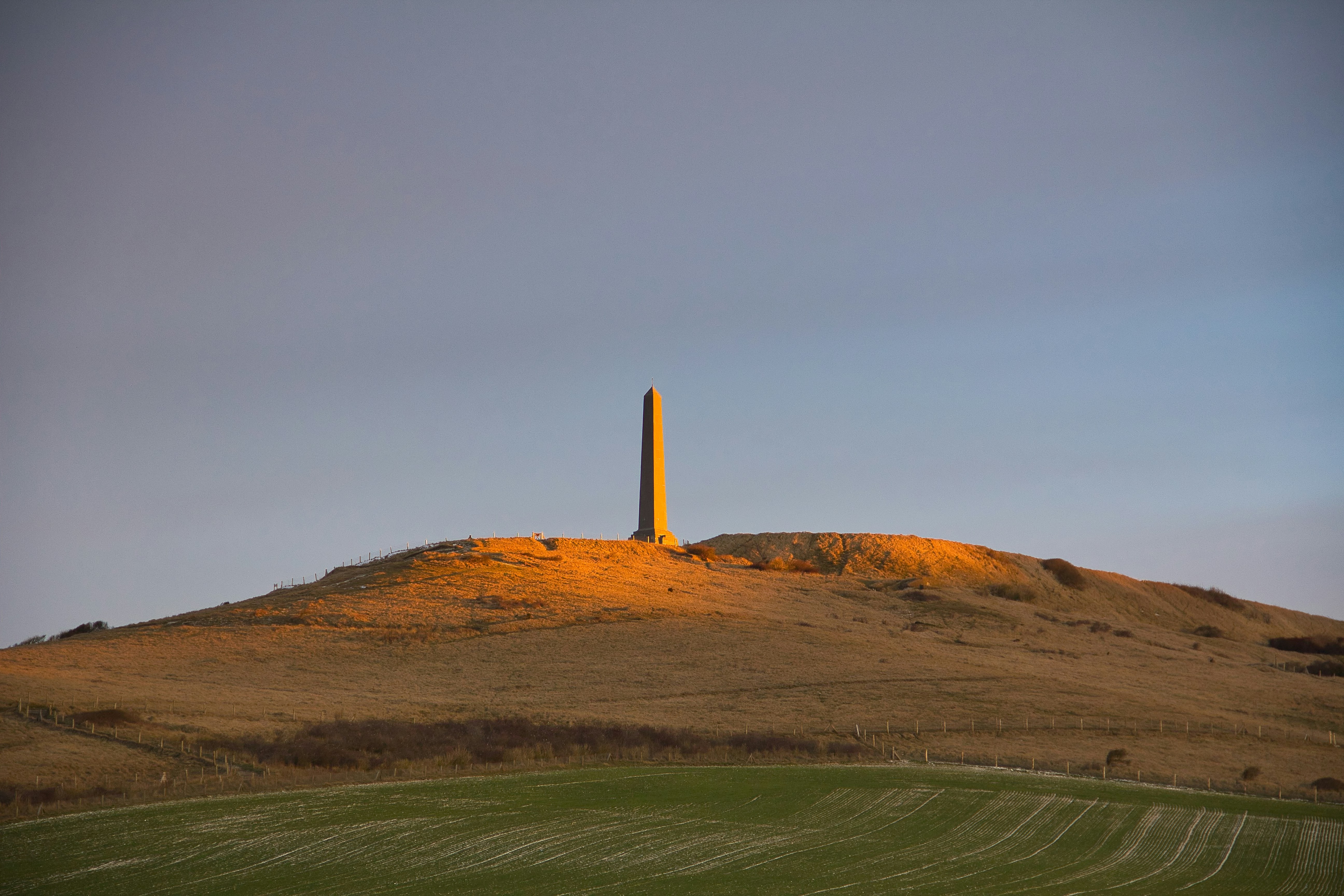 Rising sun beam striking the monolith in the early morning | brown and white tower on green grass field under blue sky during daytime