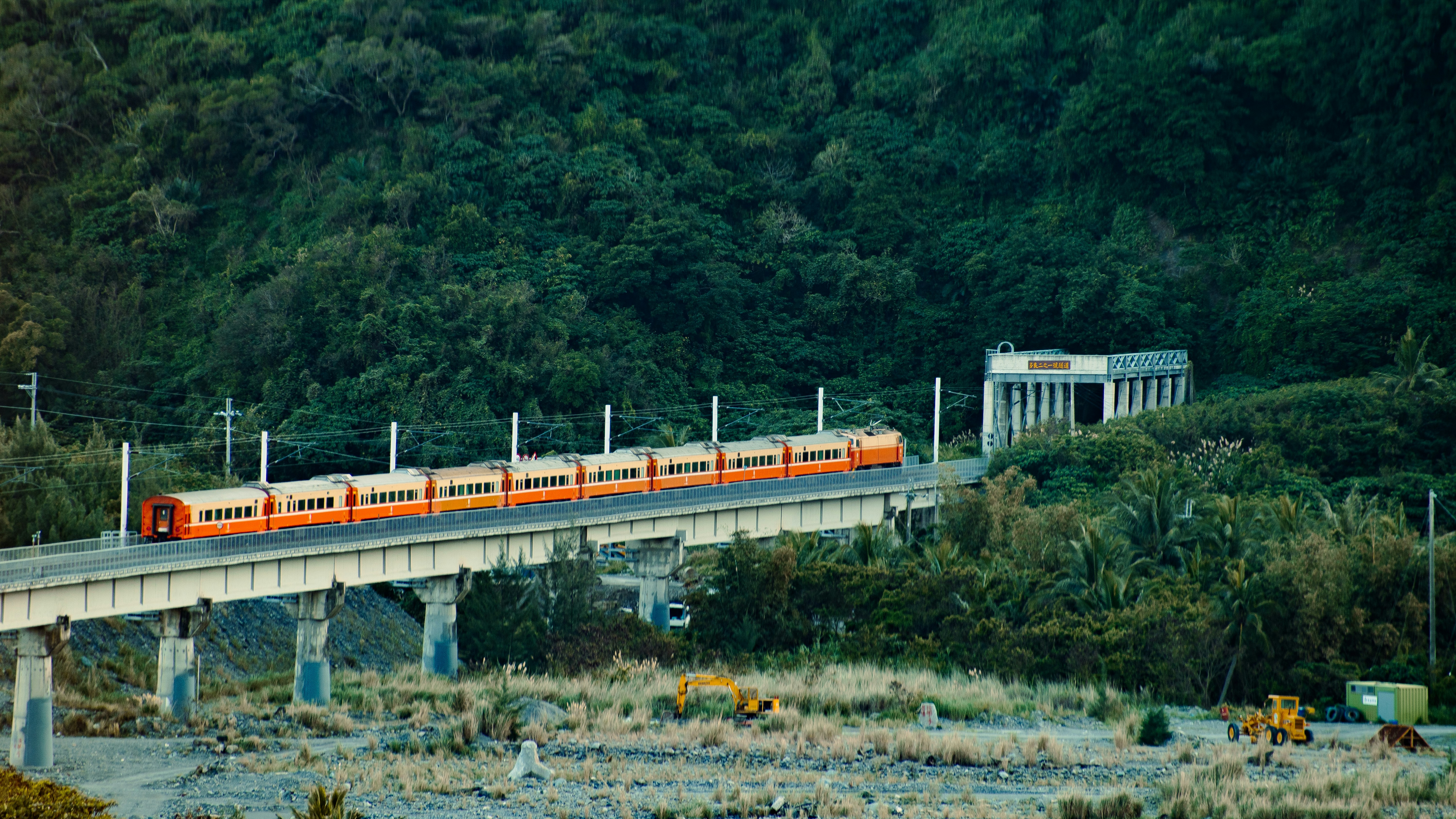 An orange train traversing a bridge, surrounded by lush greenery and distant structures, illustrating the harmony between infrastructure and nature.