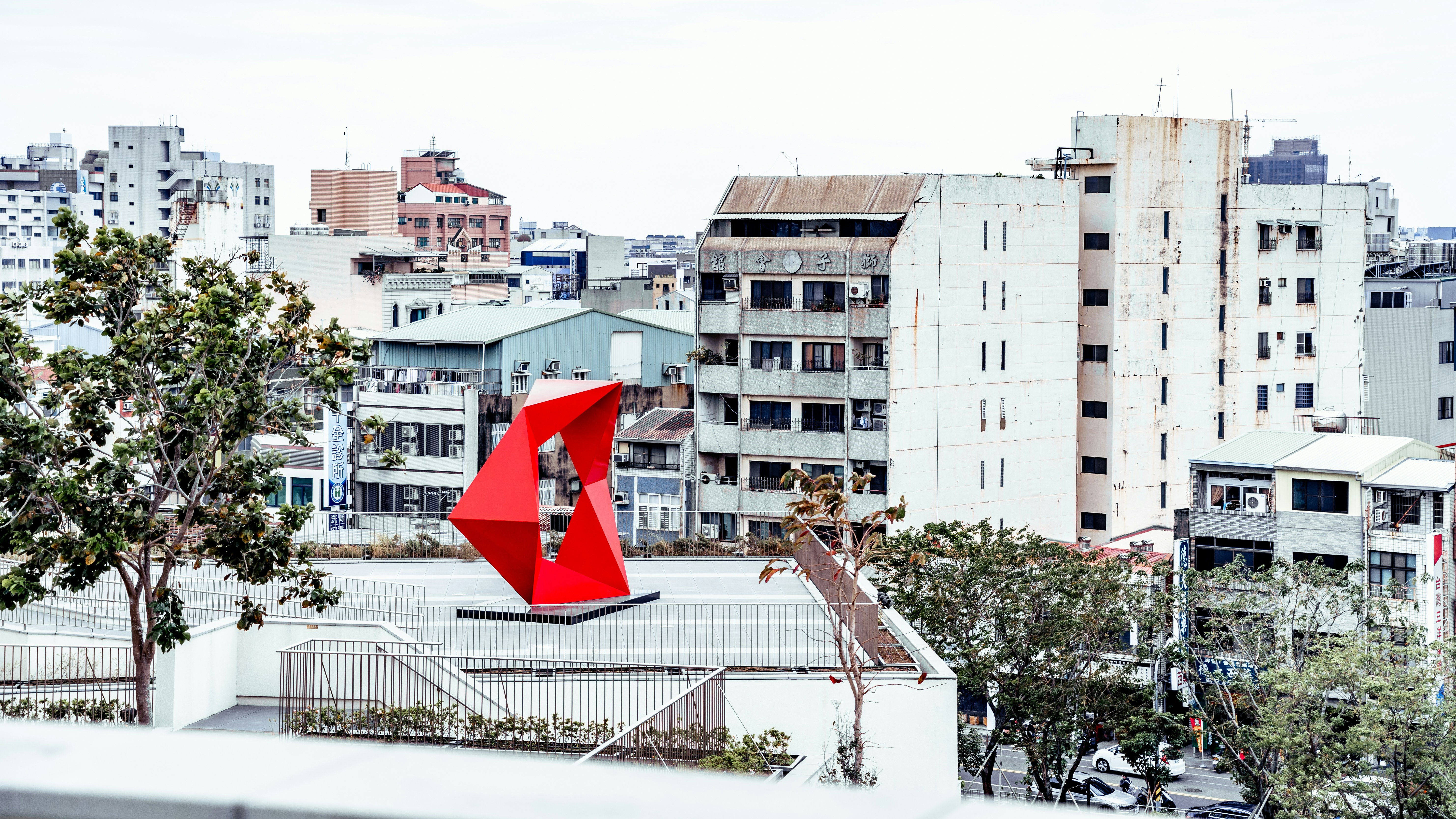 Red and white flag on a white metal fence near a concrete building, set against a backdrop of urban architecture during daytime.