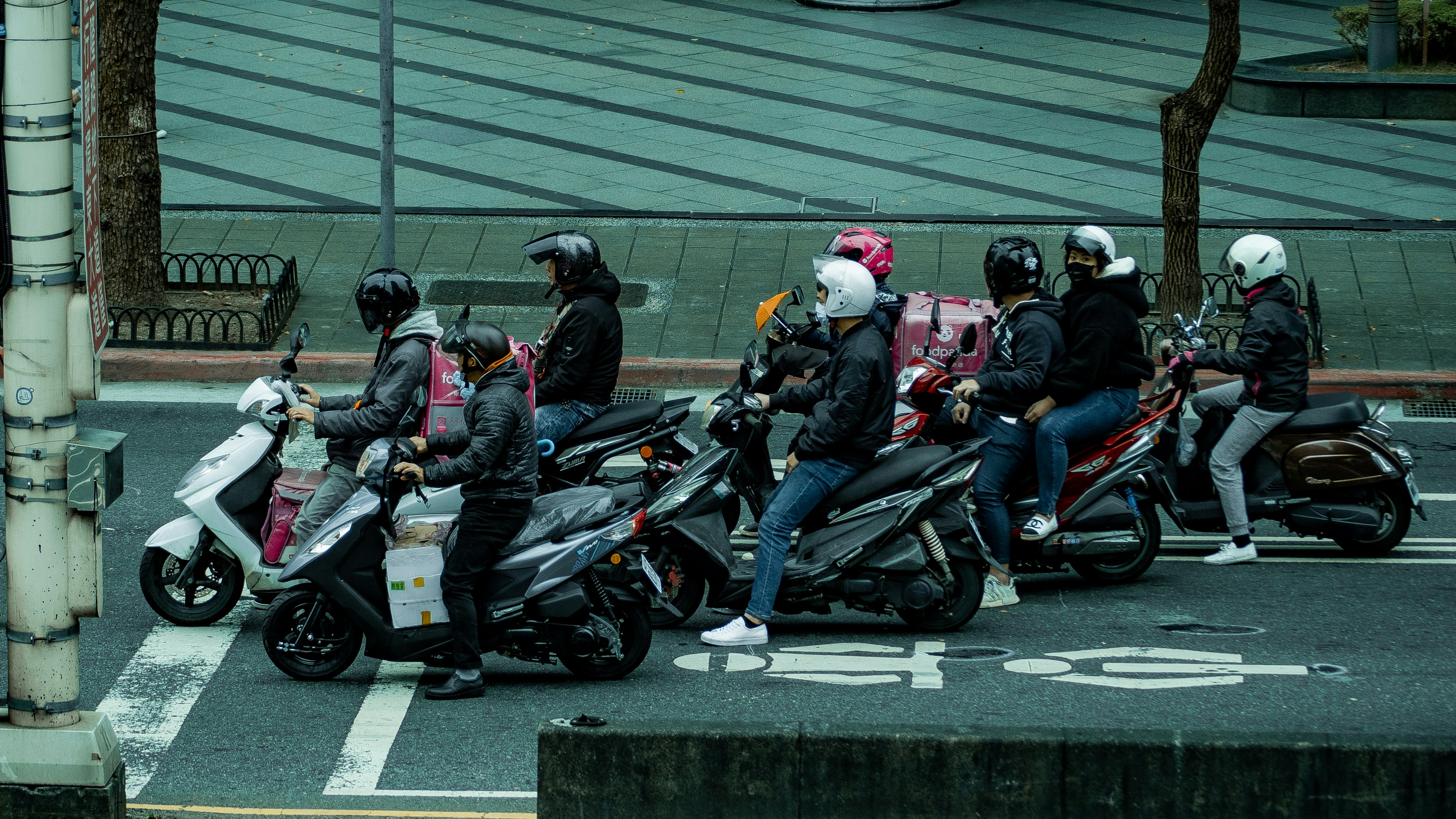 A group of delivery riders on scooters waiting at a traffic light in a bustling urban environment.