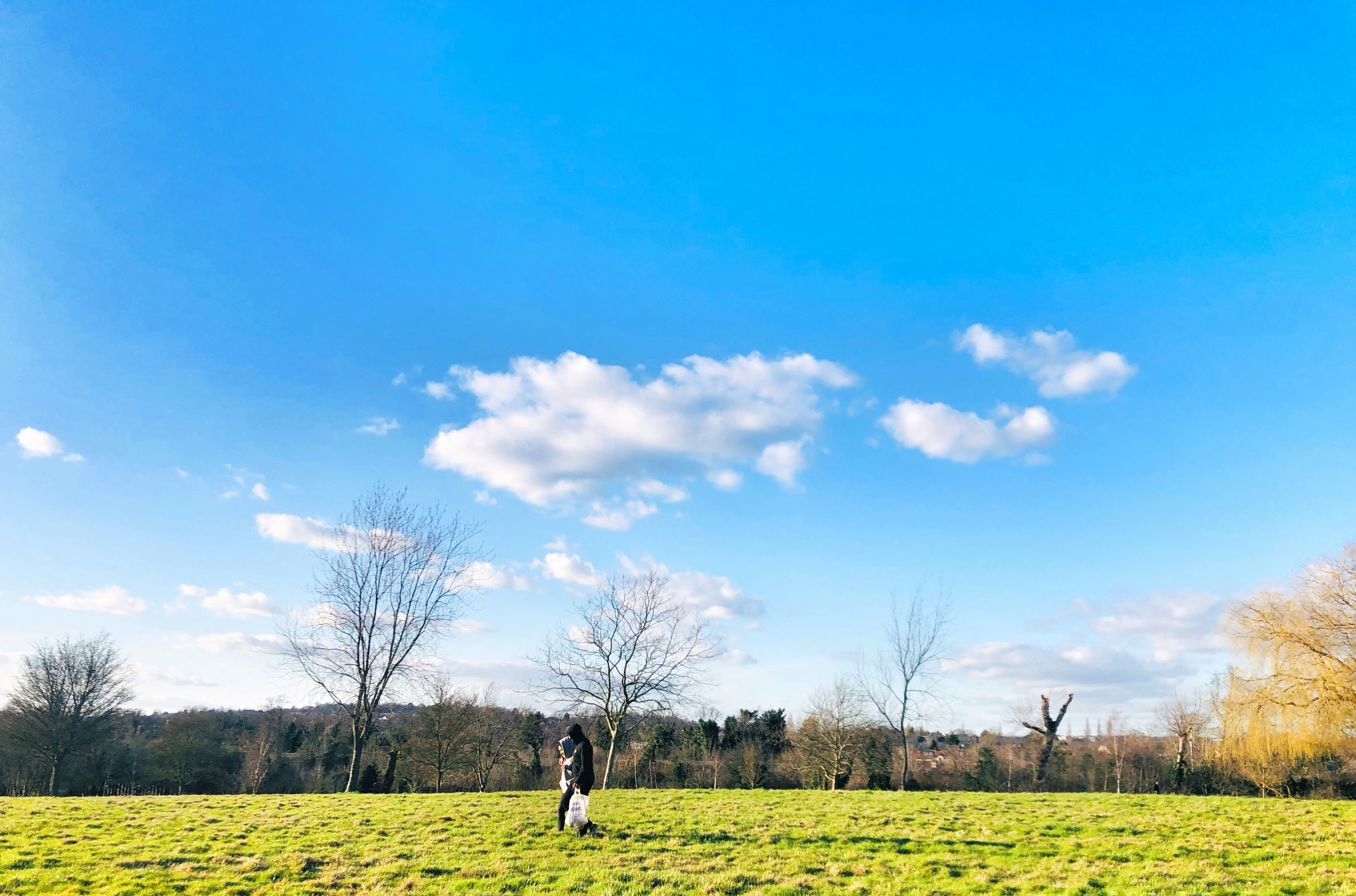 Person in a black jacket and white pants walking across a green field under a bright blue sky with scattered clouds.