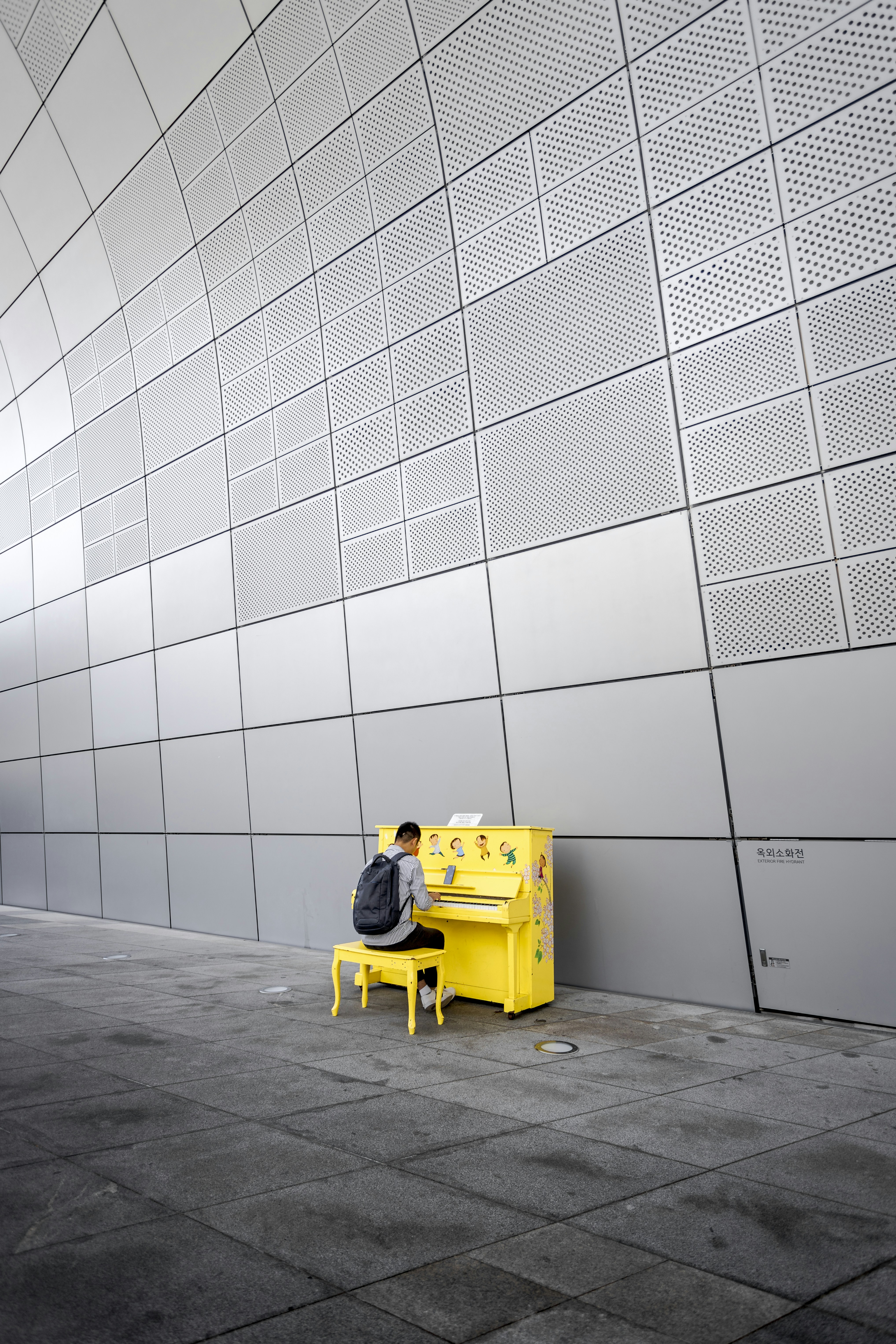 A person plays a vibrant yellow piano against a sleek, geometric gray wall, capturing a moment of creativity in an urban setting.