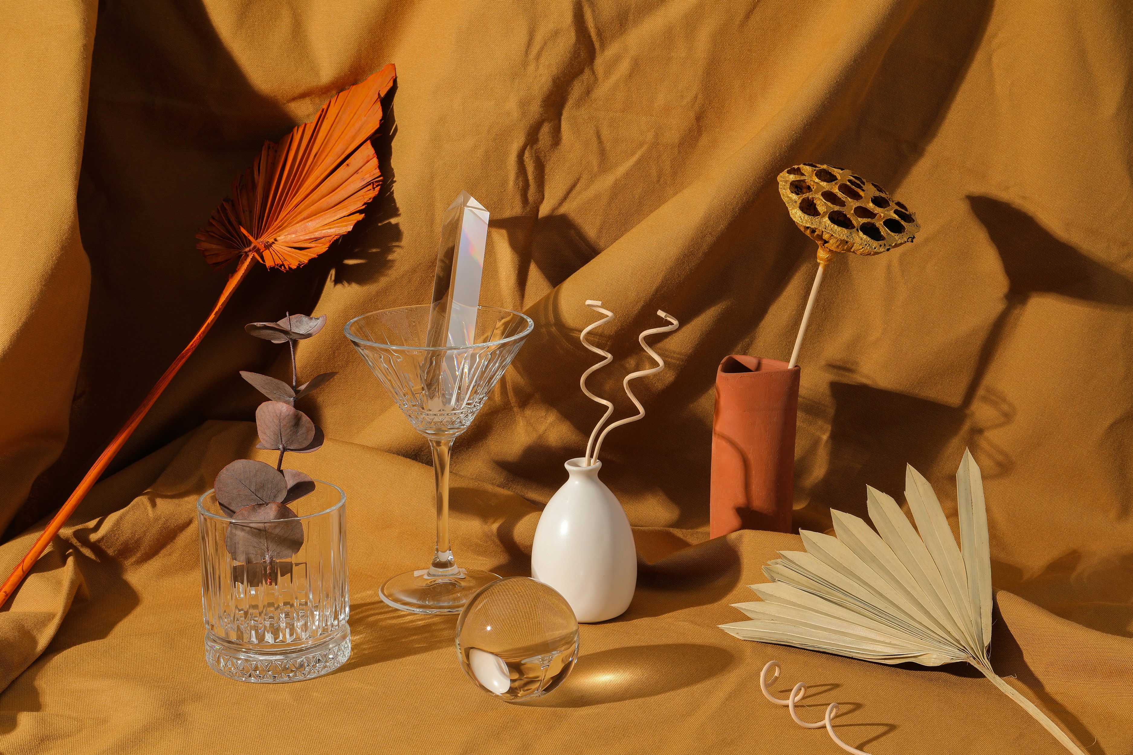 A collection of artistic objects including a crystal, glassware, and dried foliage arranged on a warm, golden fabric backdrop.