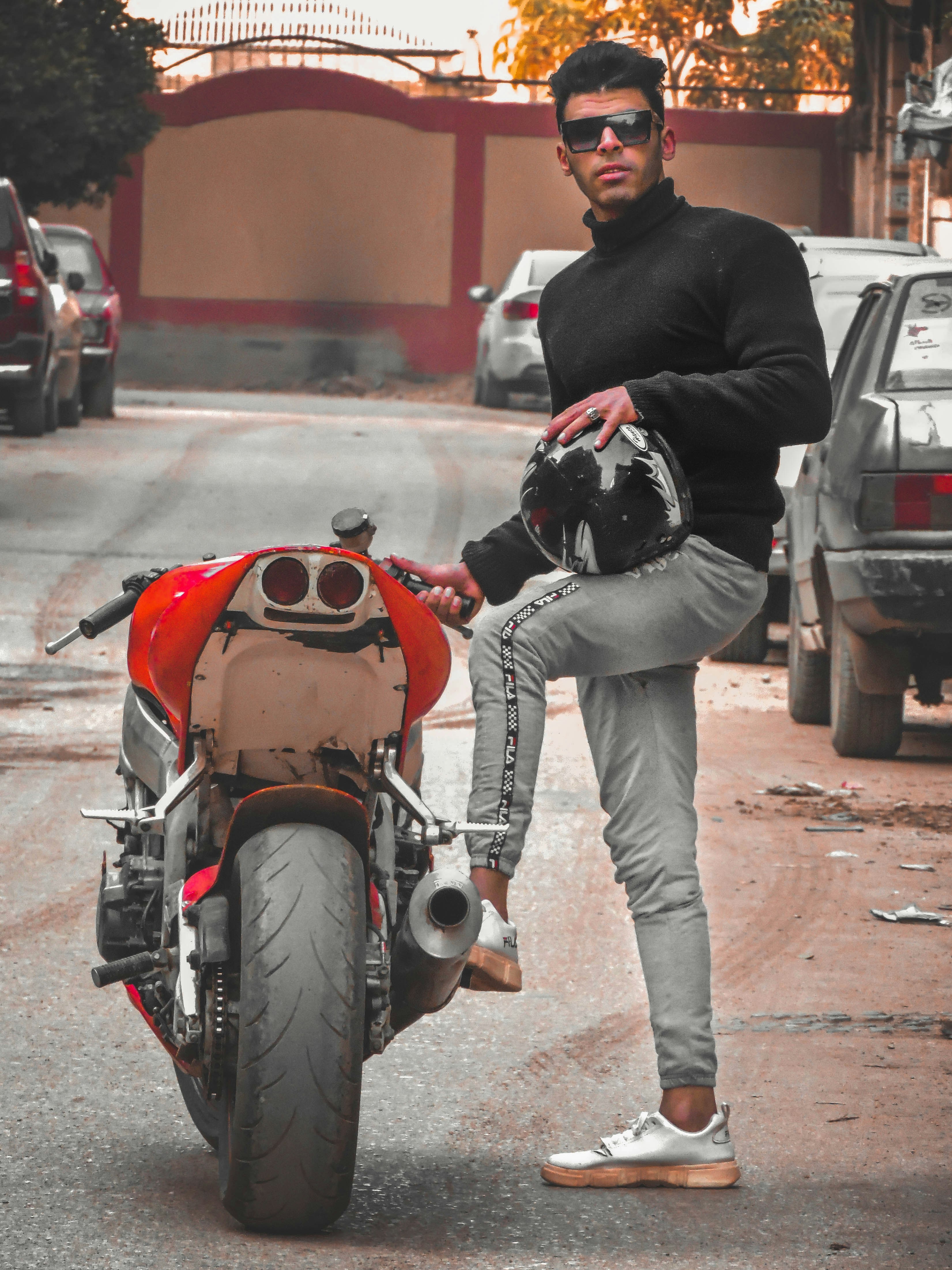 A man in a black turtleneck and sunglasses stands beside a red sport motorcycle on a gritty city street, helmet in hand.