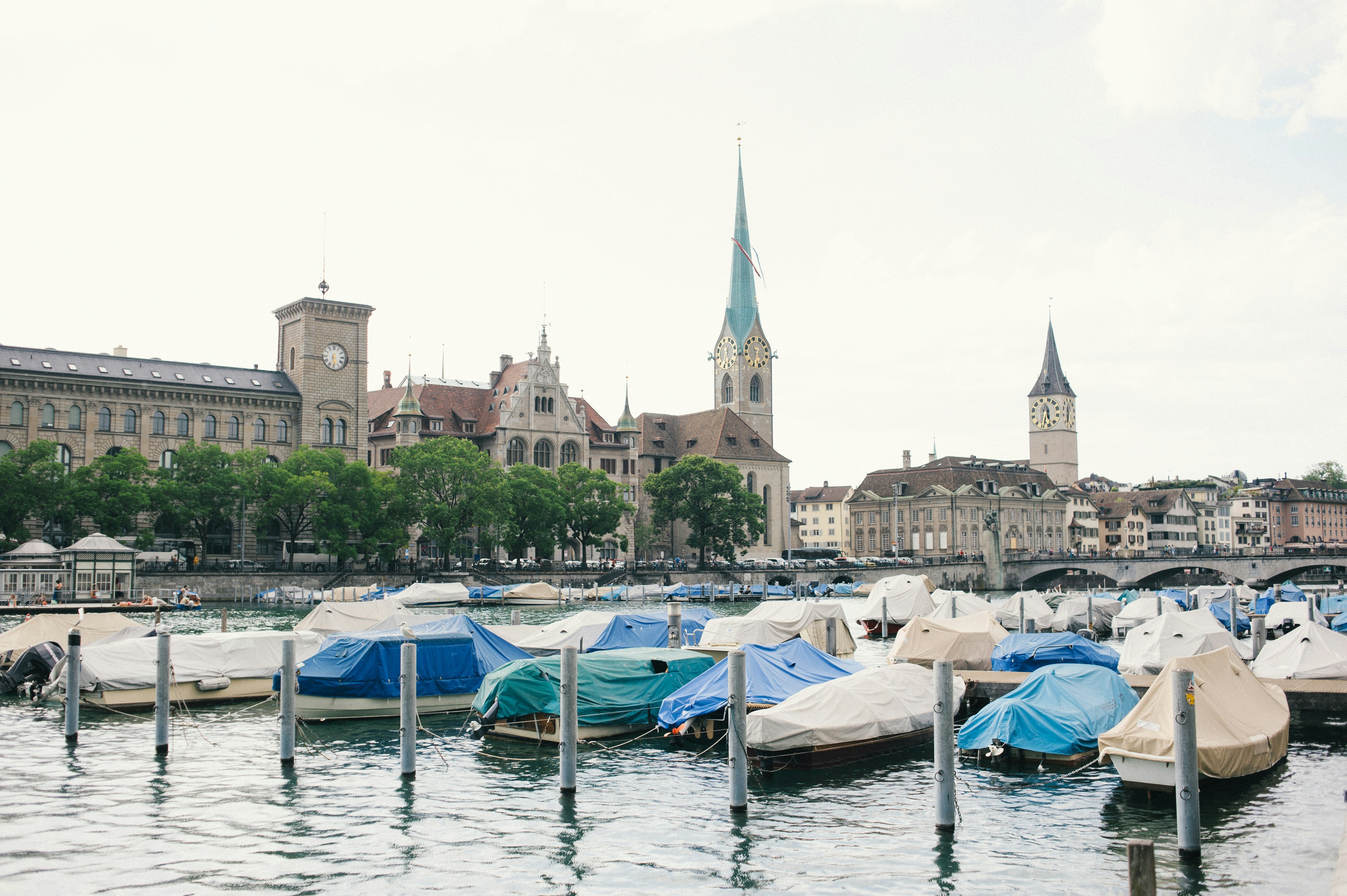 Colorful boats anchored in Zurich's harbor with historical buildings and a clock tower in the background. The scene reflects a tranquil waterfront setting.