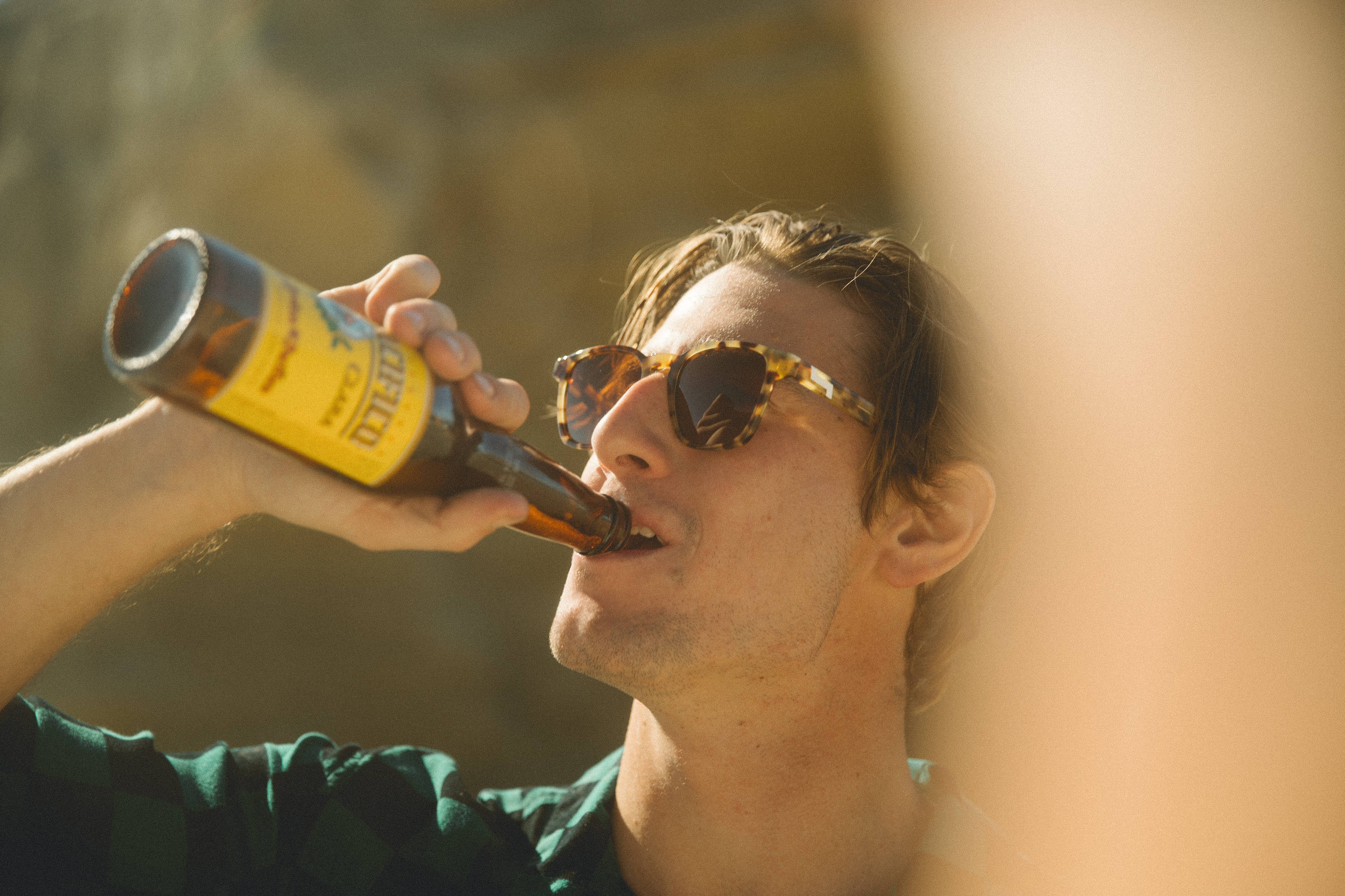 Person wearing sunglasses enjoying a drink from a bottle under warm sunlight.