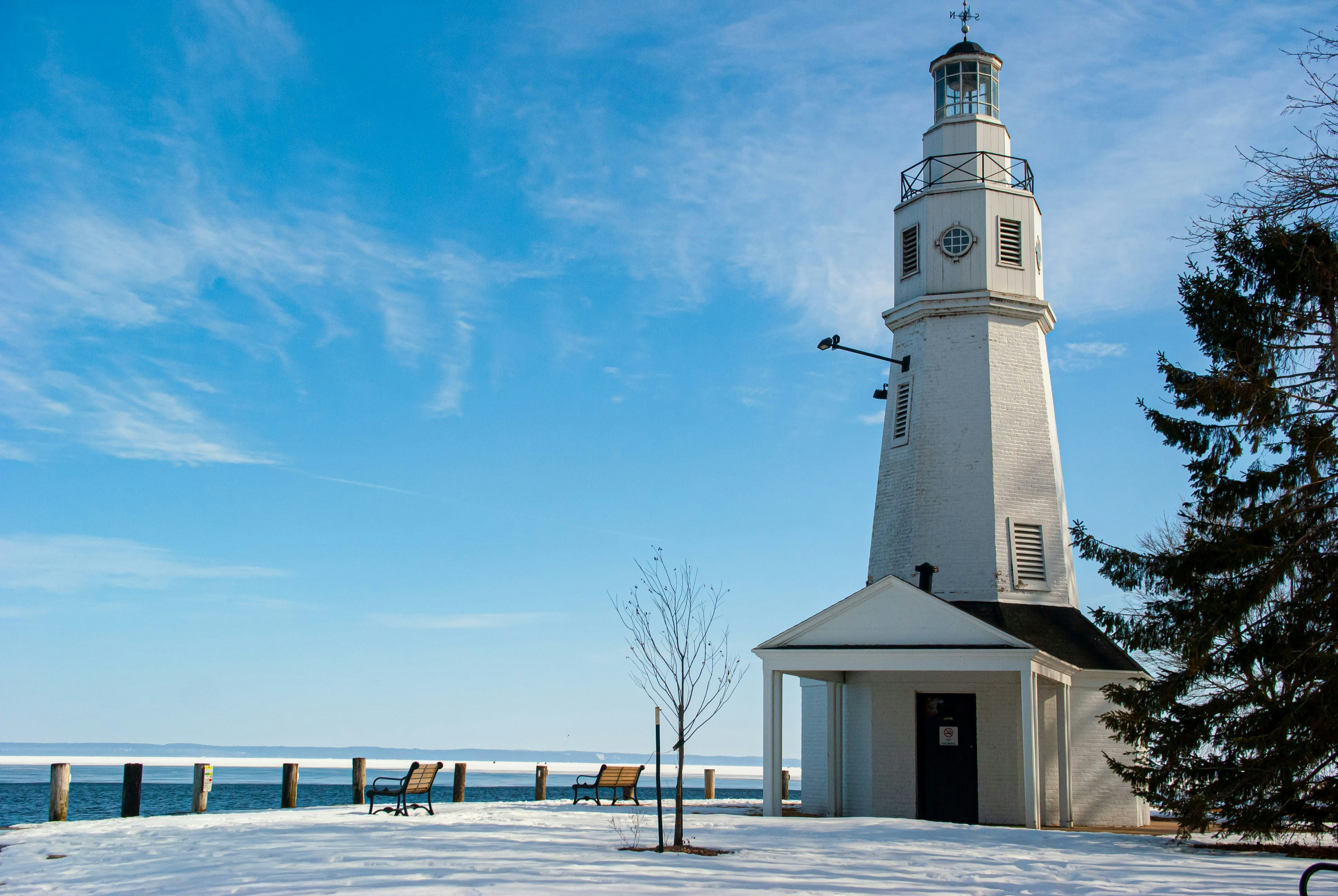 Lighthouse standing tall against a clear blue sky, surrounded by snow and a serene lake view. Benches provide a tranquil spot for reflection.