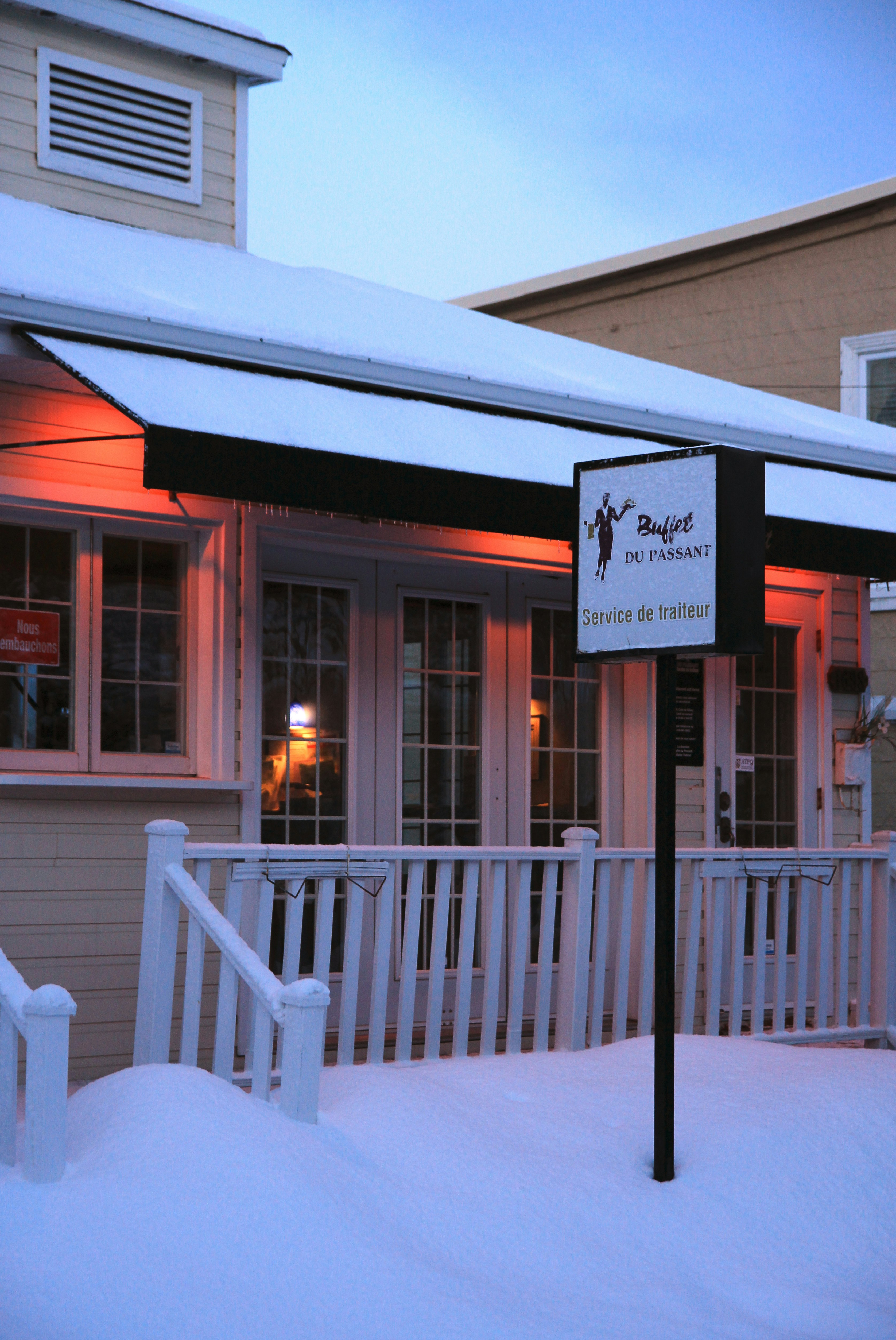 A cozy restaurant entrance adorned with snow, featuring a prominent sign and warm interior lights peeking through the windows.