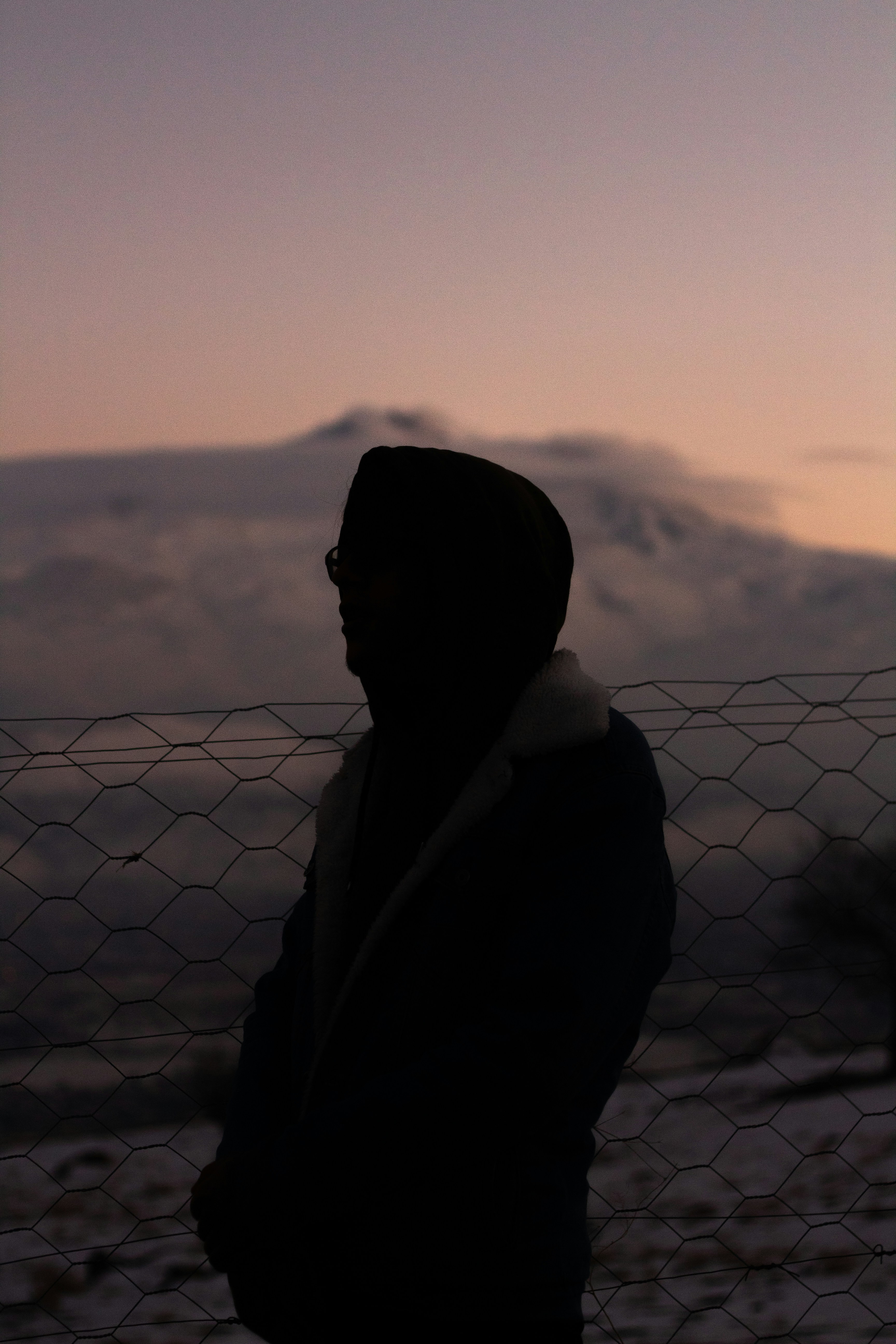 Hooded Silhouette by a Fence Against a Snowlit Dusk