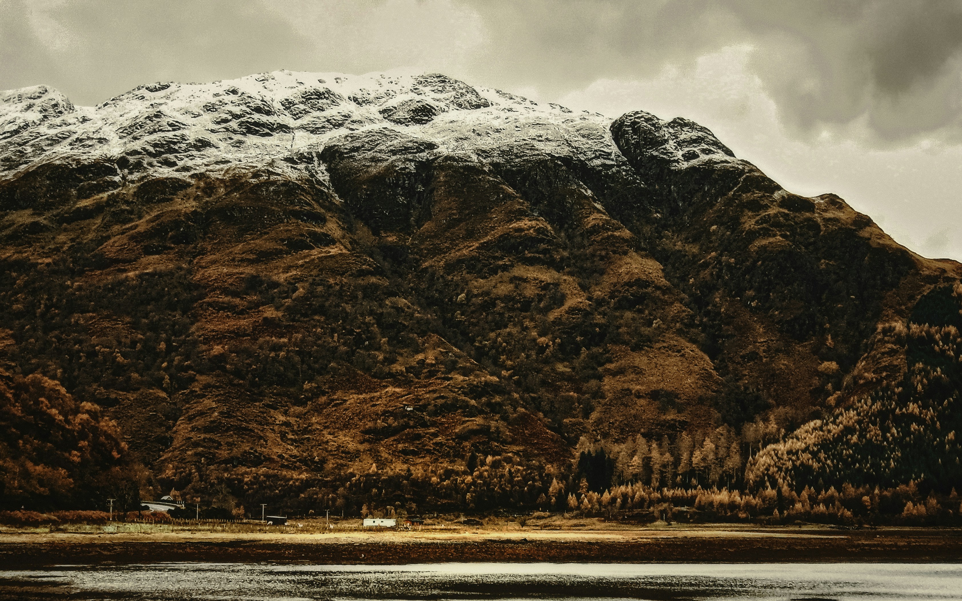 Snow-dusted mountain ridge looms over a tranquil, autumn-hued shoreline with a small cabin by the water.