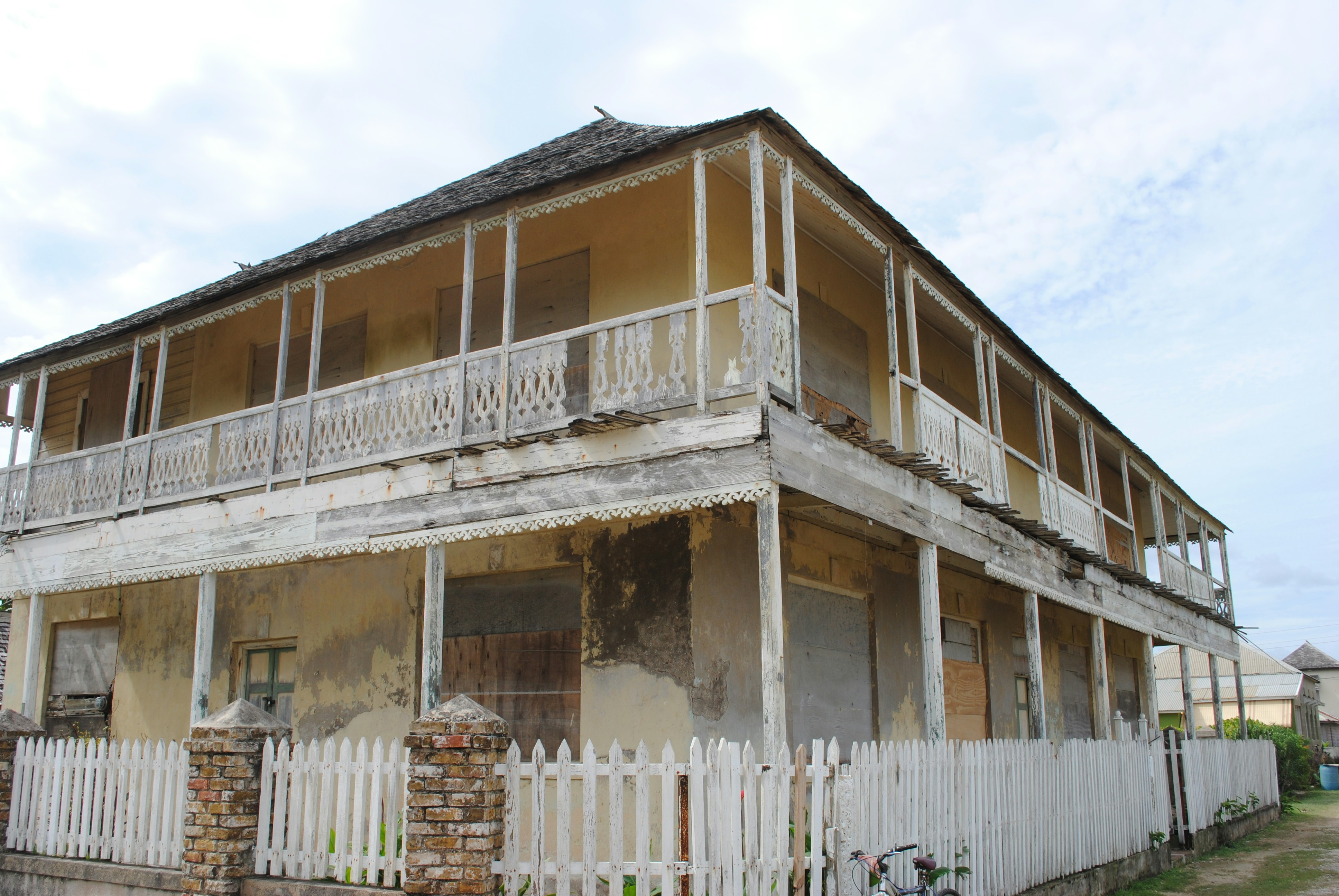 Aging two-story building with peeling paint and a white wooden fence under a cloudy sky.