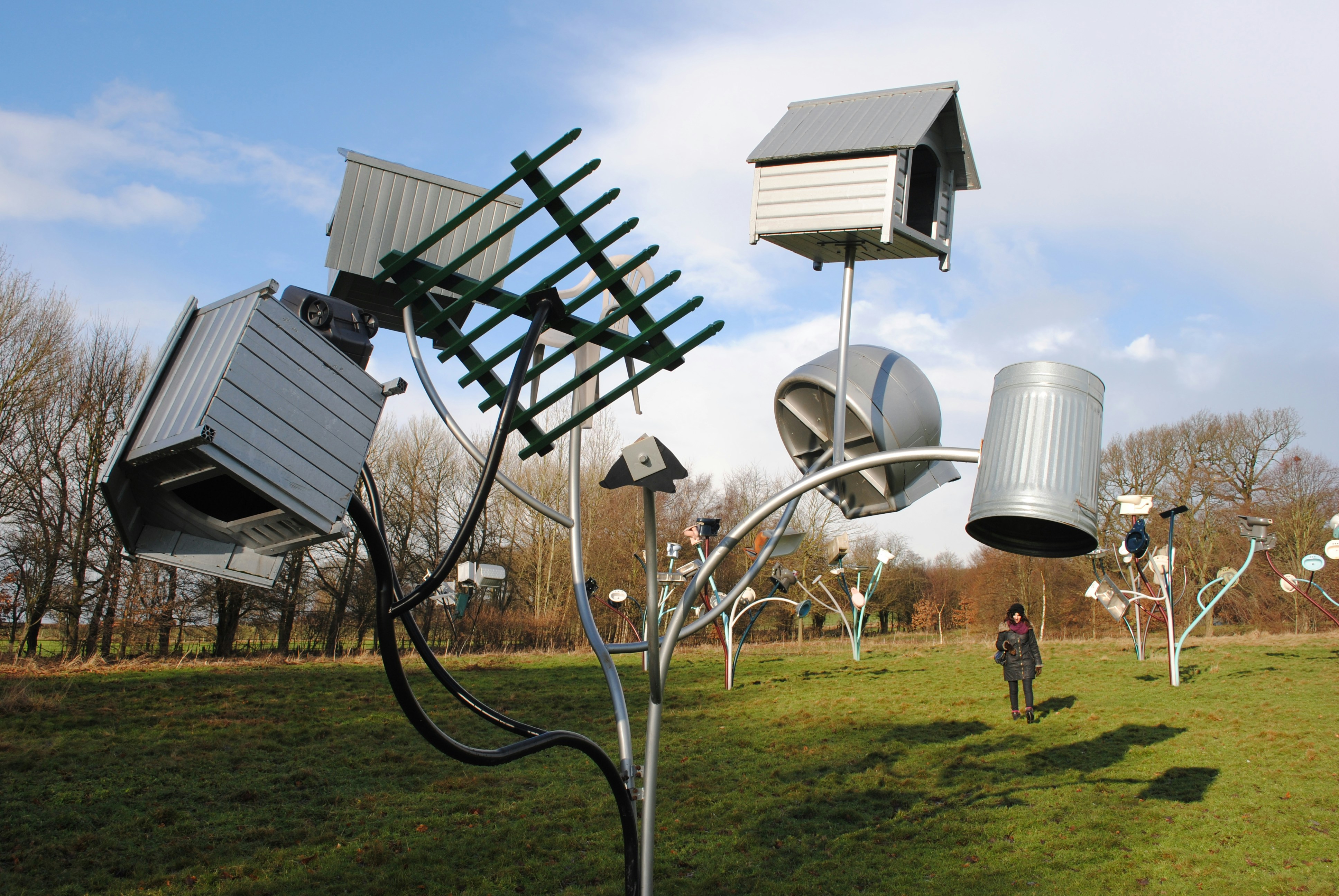 Metal tree sculpture adorned with household objects under a partly cloudy sky in a grassy field.