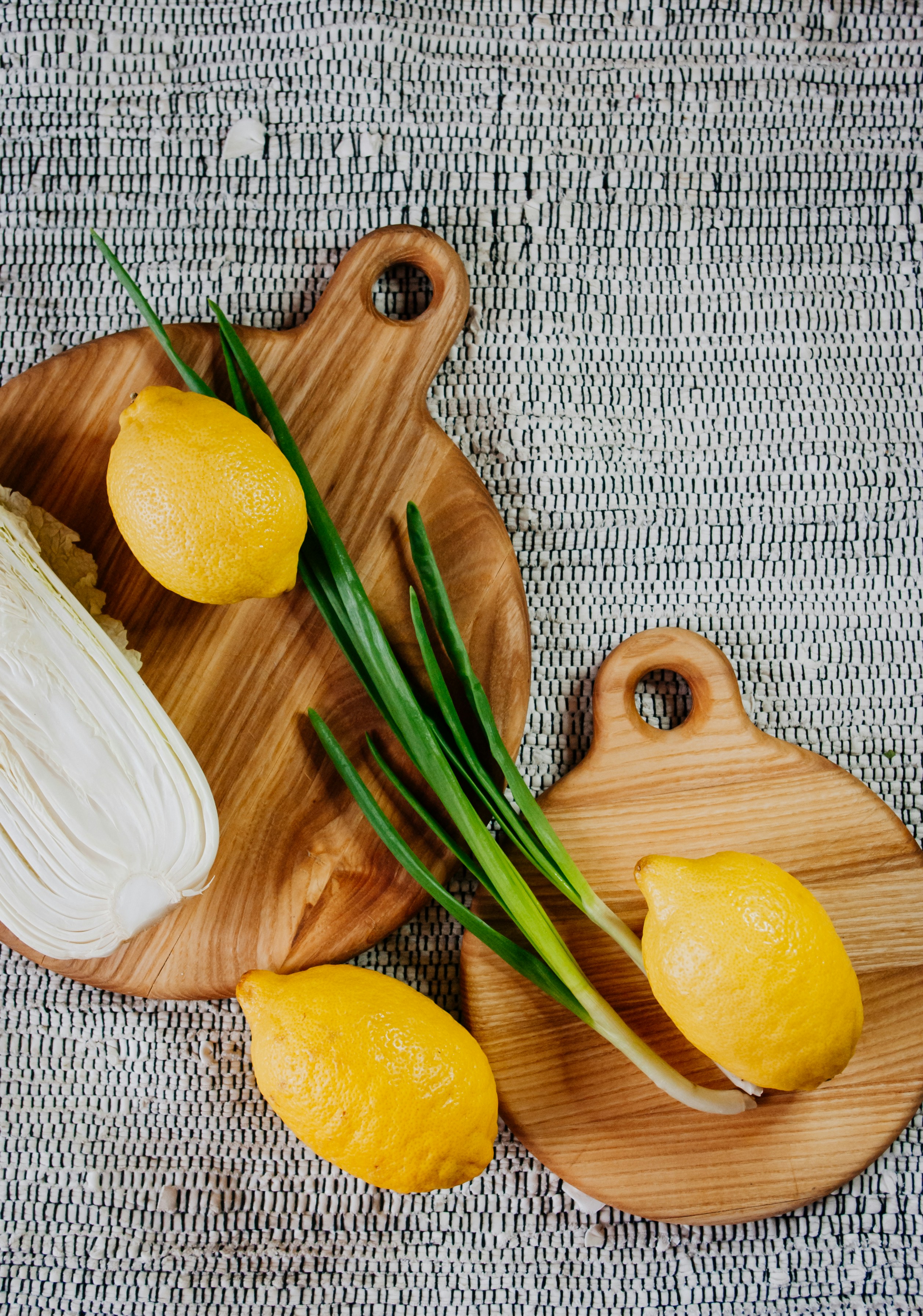 Cluster of bright lemons resting on round wooden boards with green onion shoots on a textured fabric background. A still-life setup emphasizing fresh citrus and rustic serving boards.