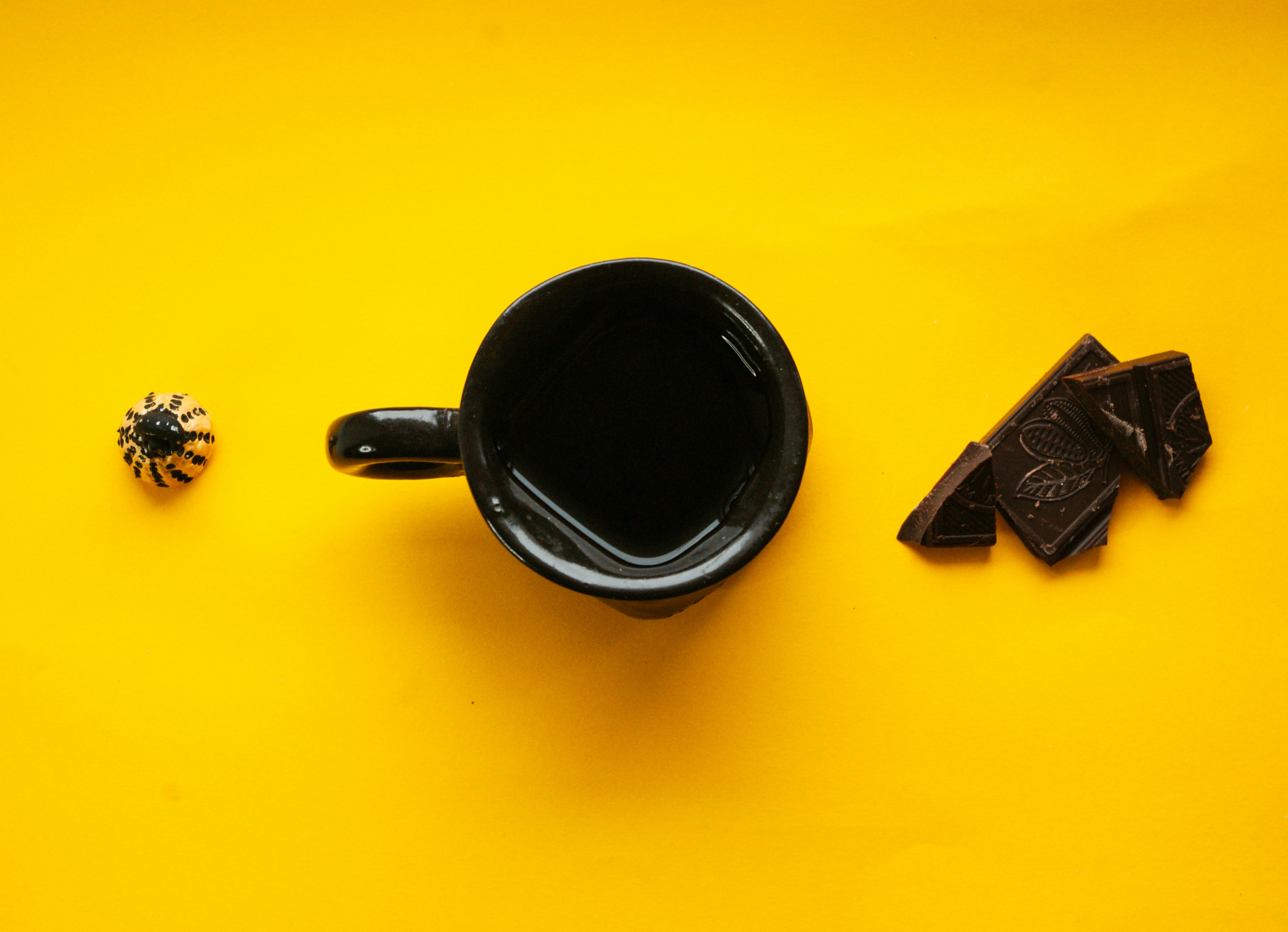 A black coffee mug sits on a bright yellow surface with broken chocolate pieces to the right and a small confection to the left, photographed from a top-down perspective.