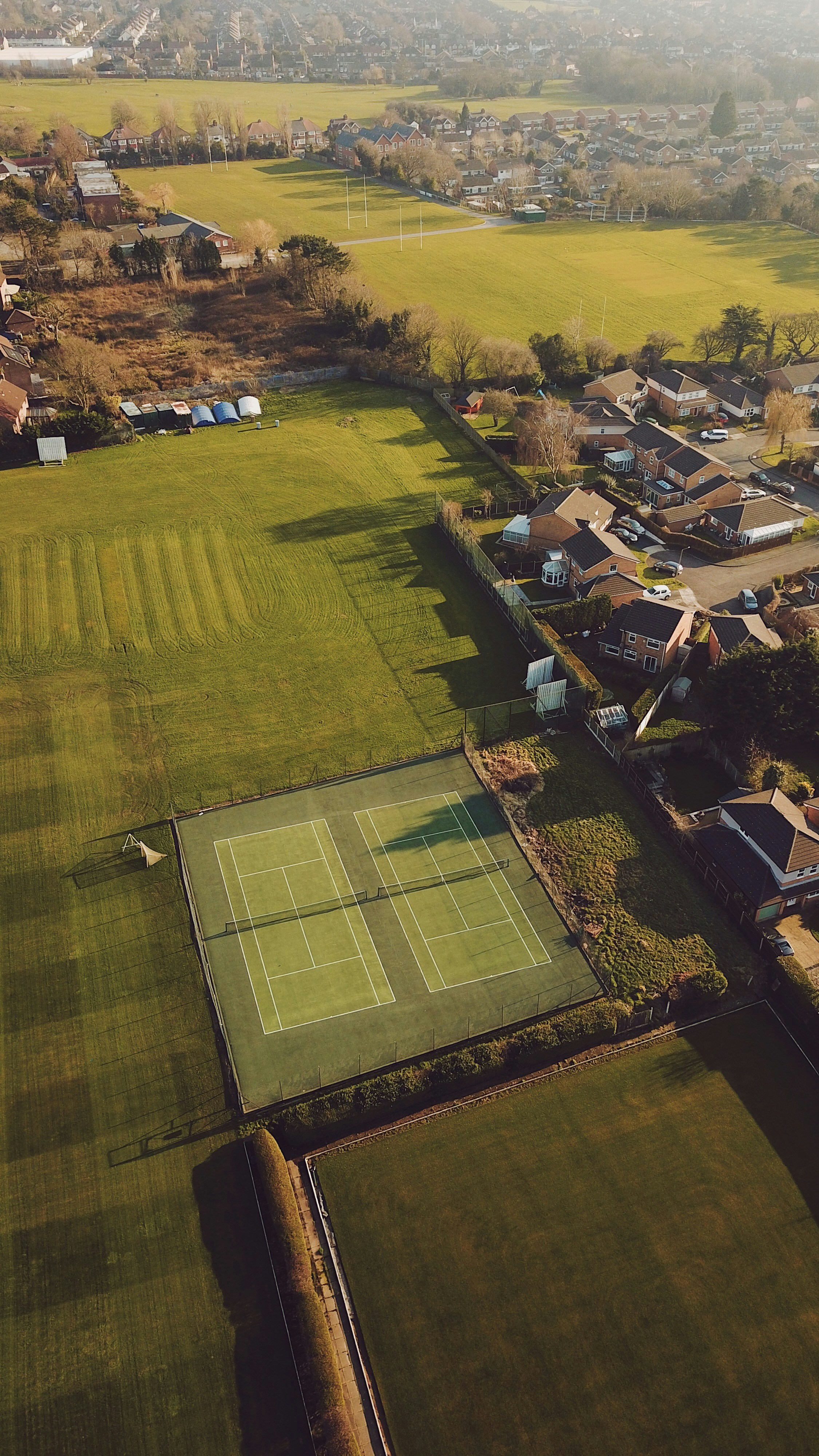 Aerial view showcasing a tennis court surrounded by lush green fields and residential areas, highlighting the blend of sports and community living.