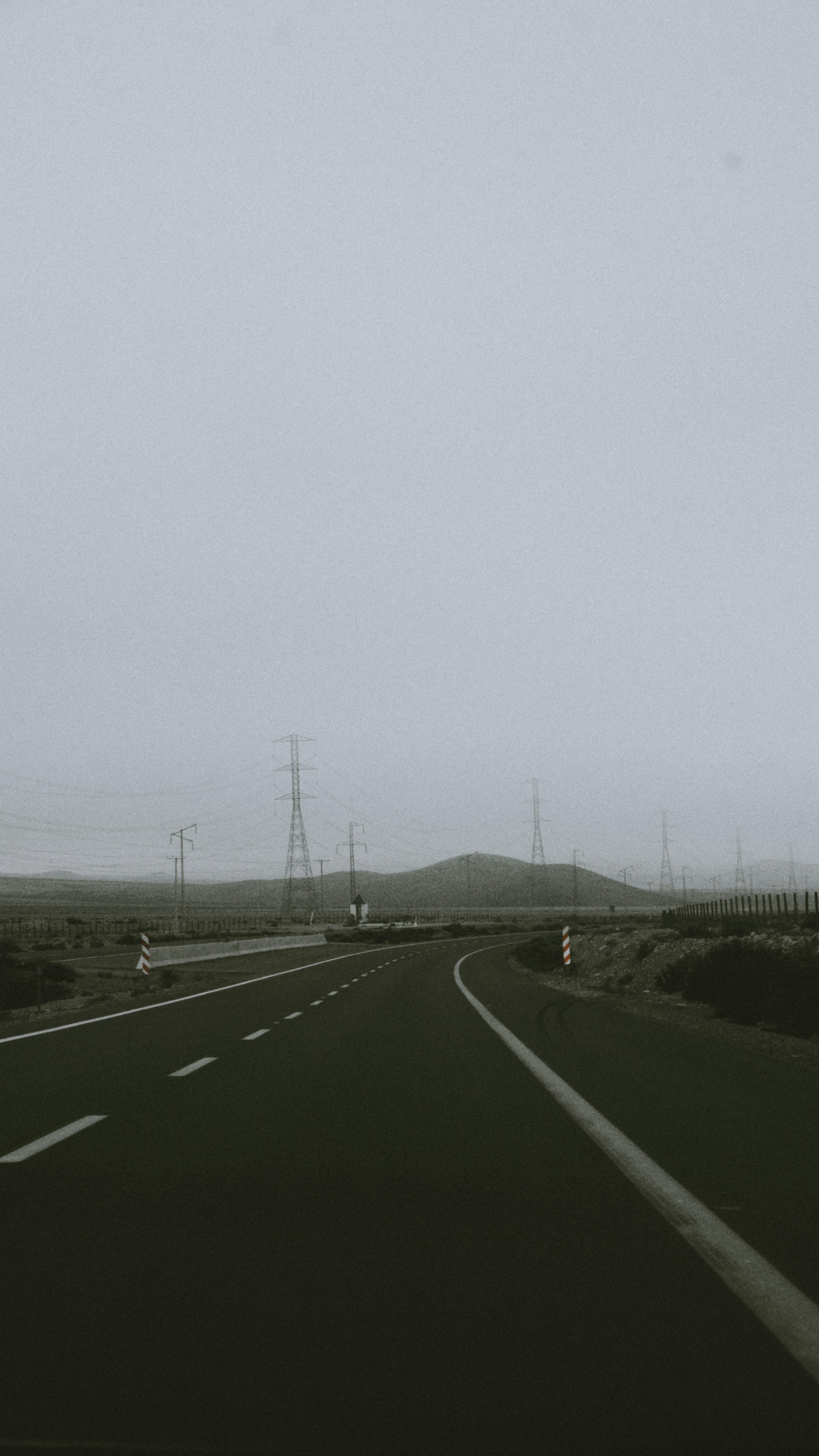 Curving road under a muted sky, flanked by distant hills and power lines, evoking a sense of solitude and journey.