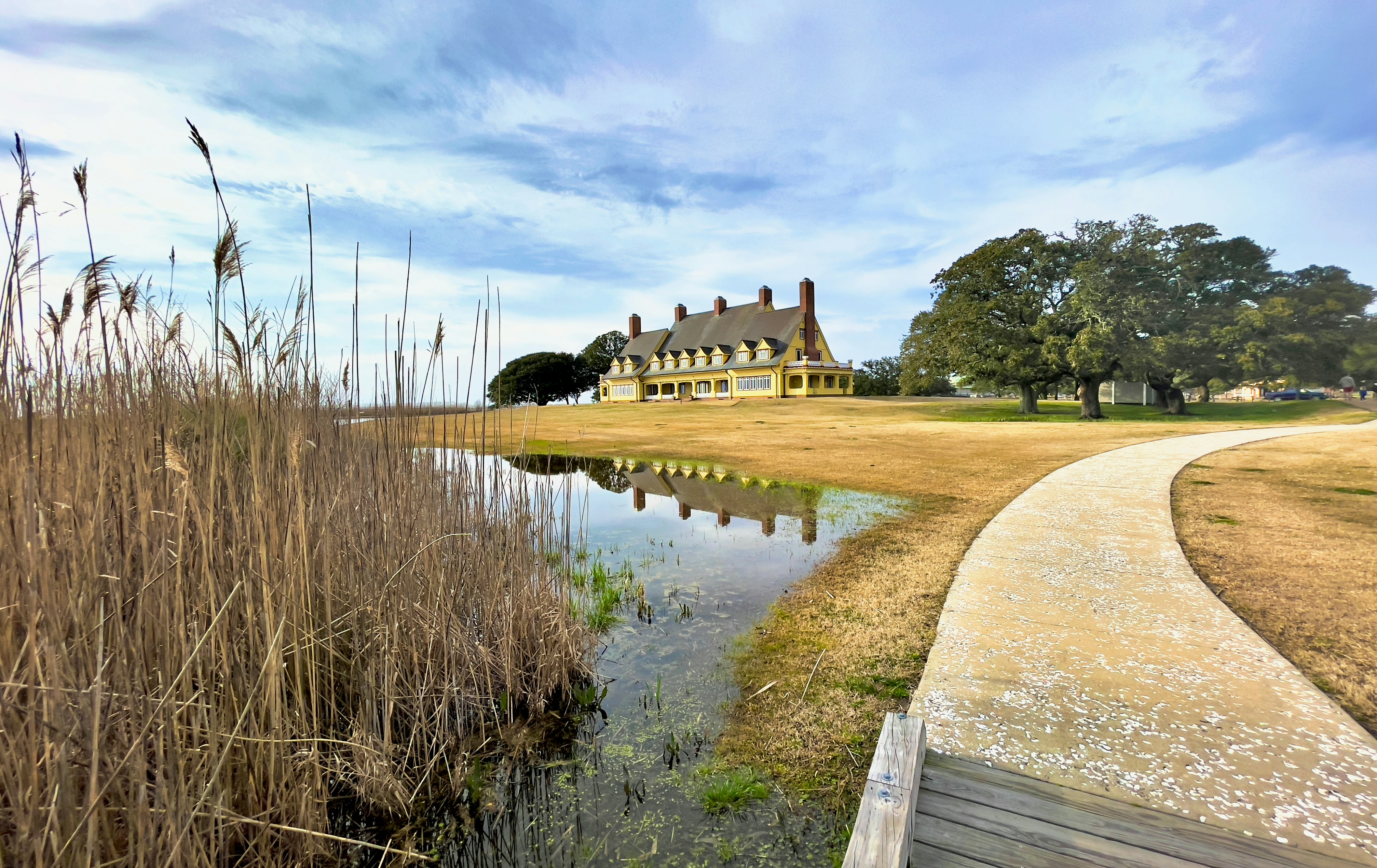 Historic house beside a calm pond, framed by tall grasses and a winding pathway. The serene landscape invites contemplation.