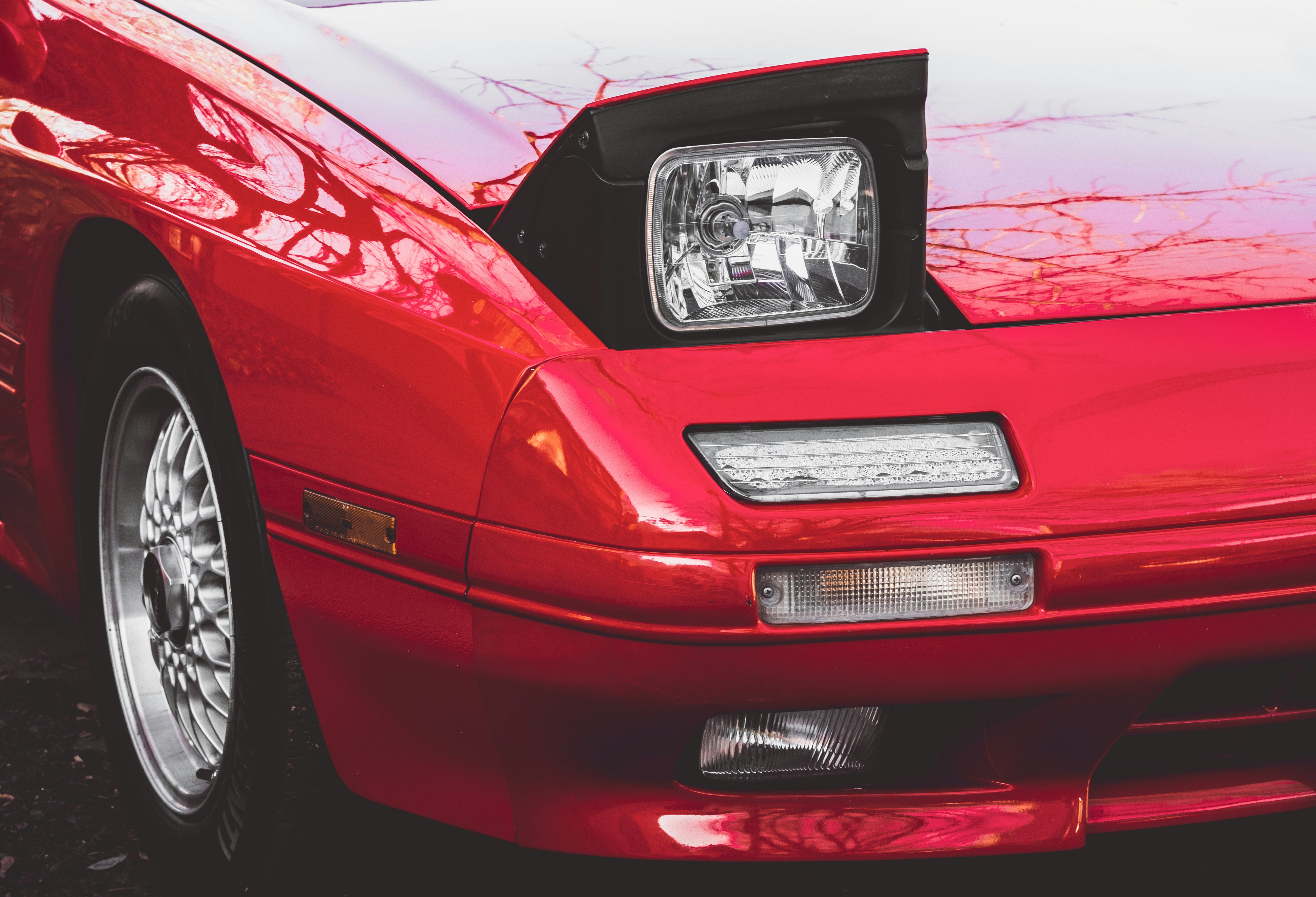 Close-up of a red sports car's pop-up headlight and sleek design with tree reflections on the hood.
