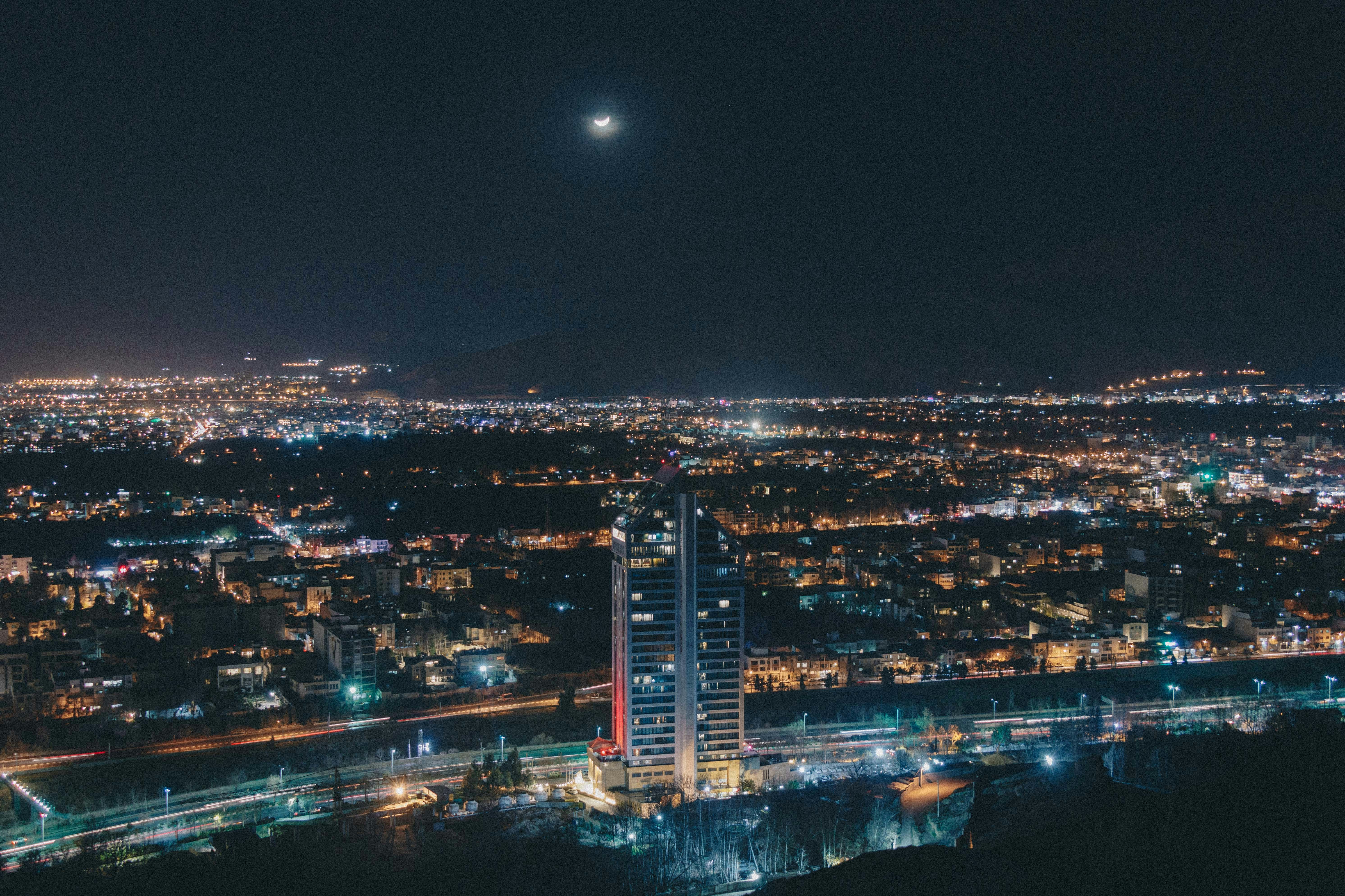 A panoramic view of a city illuminated by countless lights, with a prominent skyscraper standing tall against the night sky, highlighted by a crescent moon above.