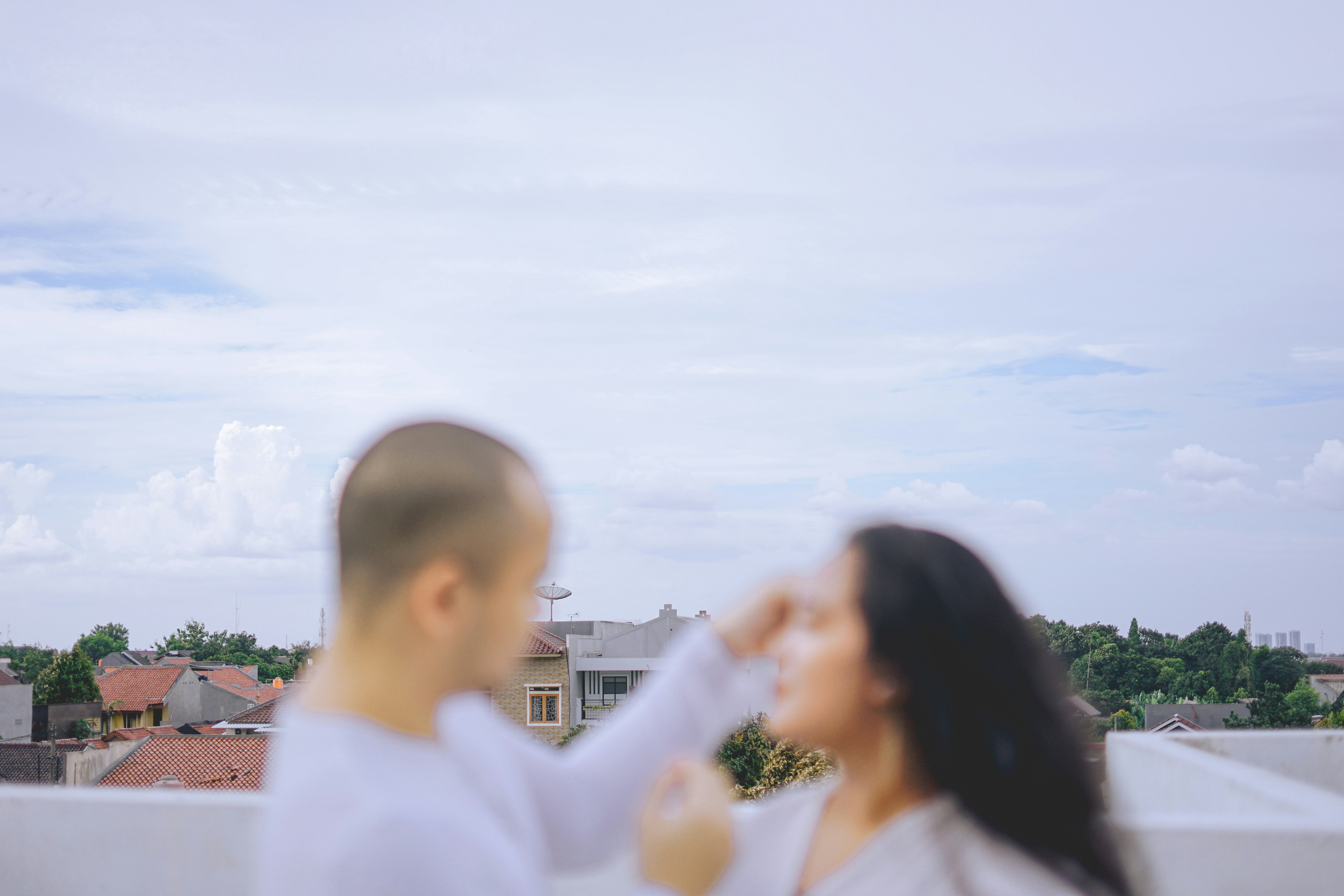Couple engaged in an intimate moment on a rooftop, with a blurred background showcasing a city skyline and soft clouds above.