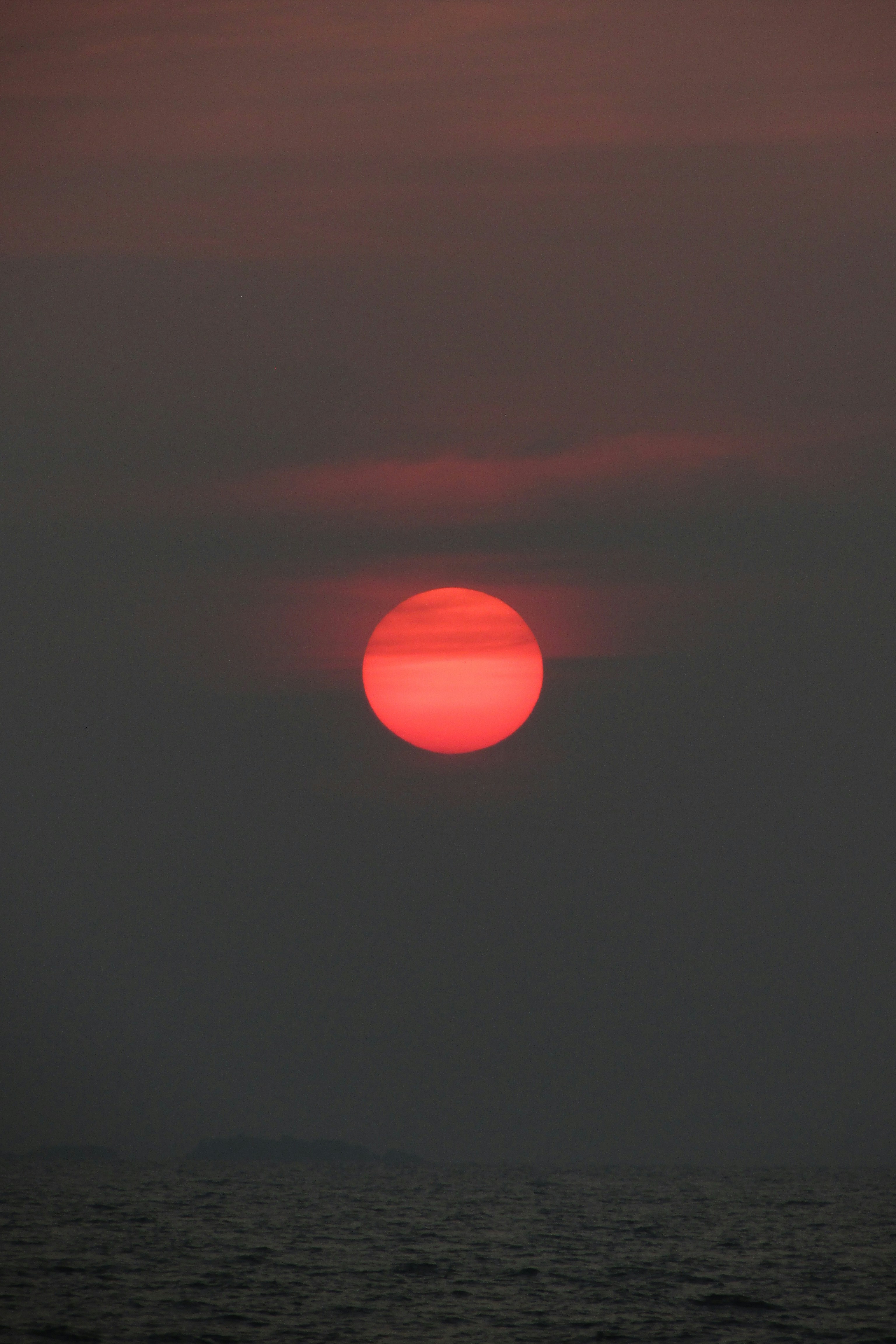Vibrant red sun setting against a hazy sky, casting a warm glow over the tranquil sea. The scene evokes a sense of calm and reflection.