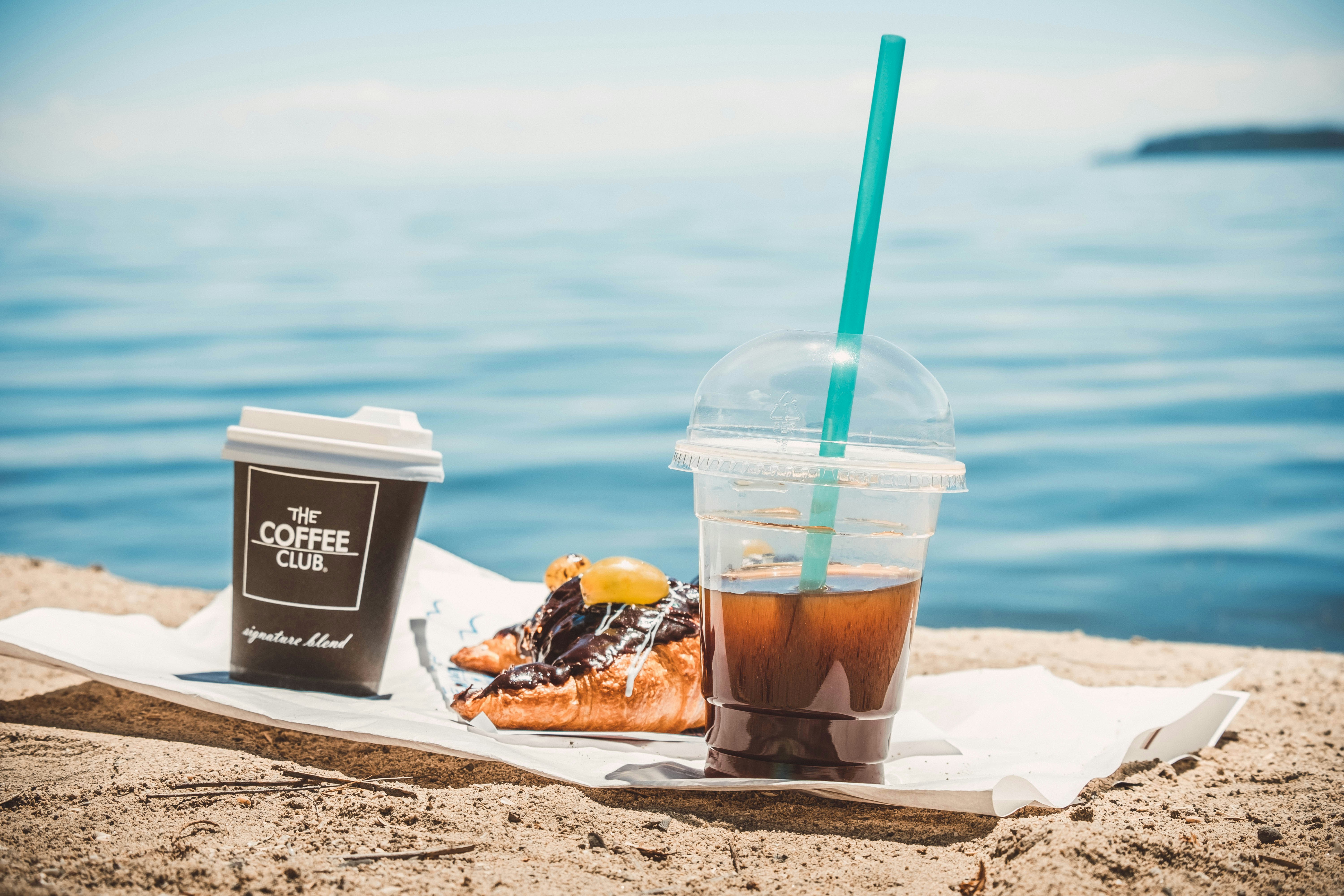 Sunlit Seaside Snack: Coffee Cup, Pastry, and Iced Drink by a Calm Blue Sea