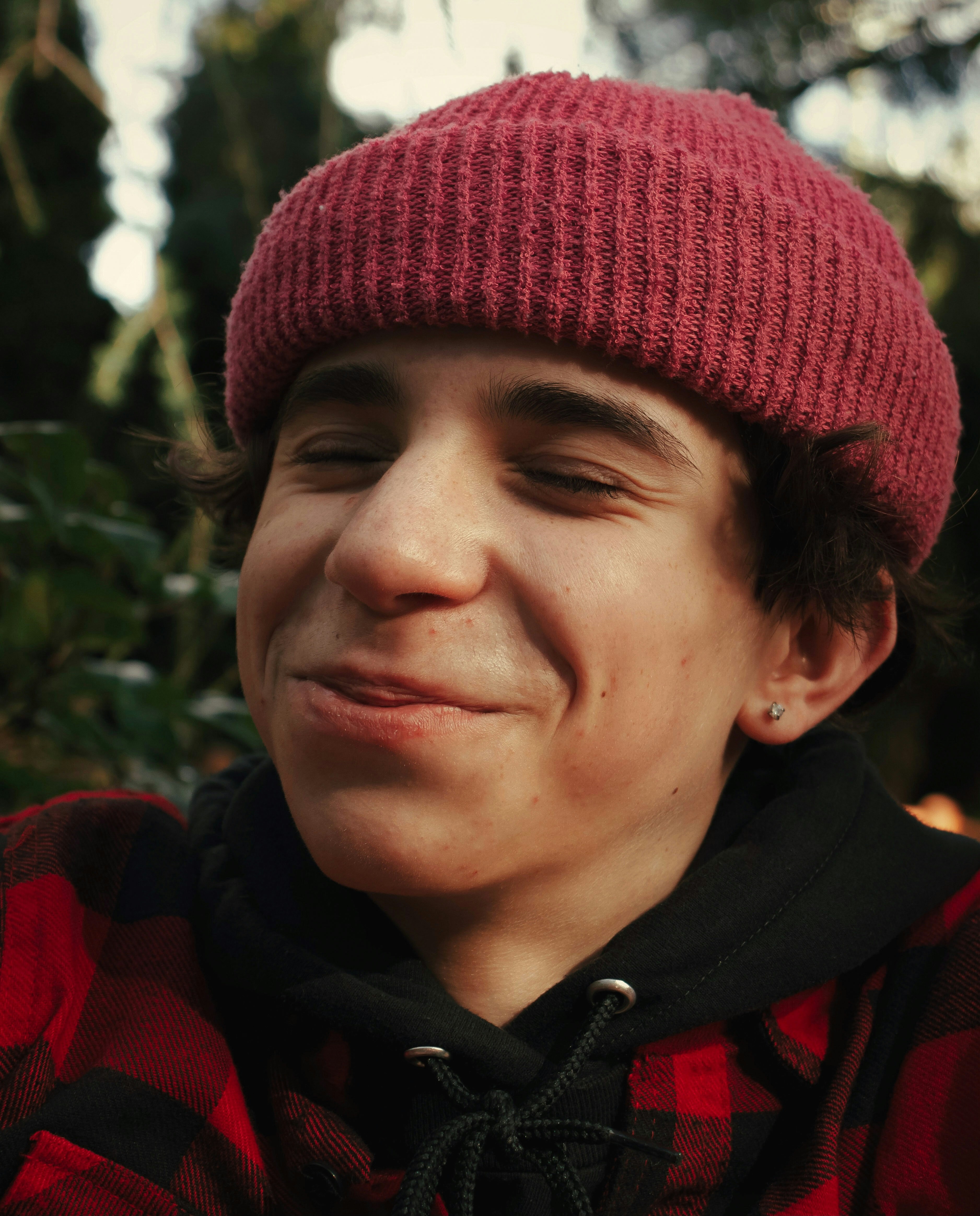 A young person with a pink beanie and plaid shirt smiles warmly, surrounded by greenery. The natural setting enhances the cheerful expression.