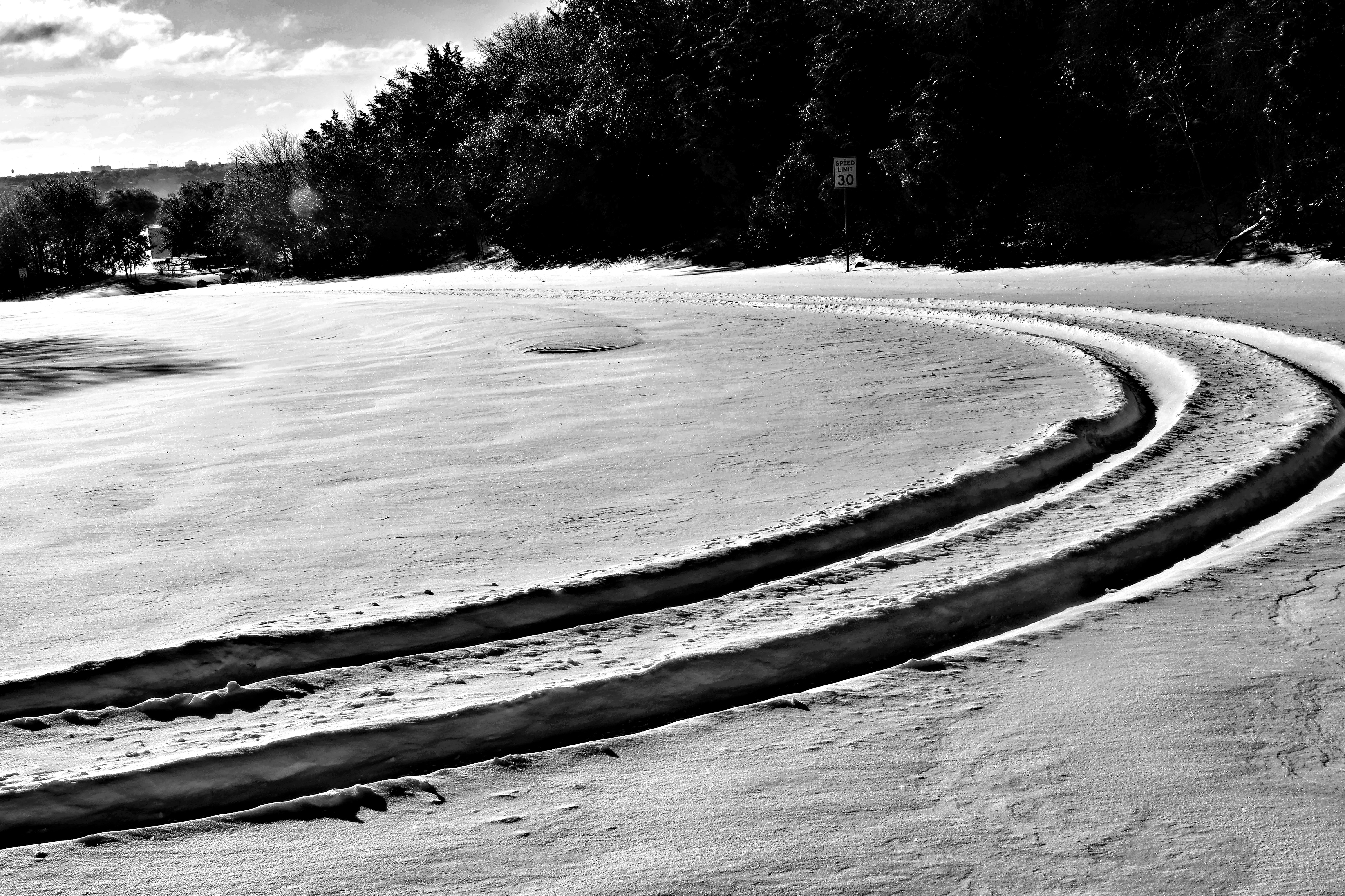 Tire tracks winding through a snow-covered landscape, revealing the serene stillness of a winter day. The monochrome palette enhances the texture of the snow and the depth of the scene.