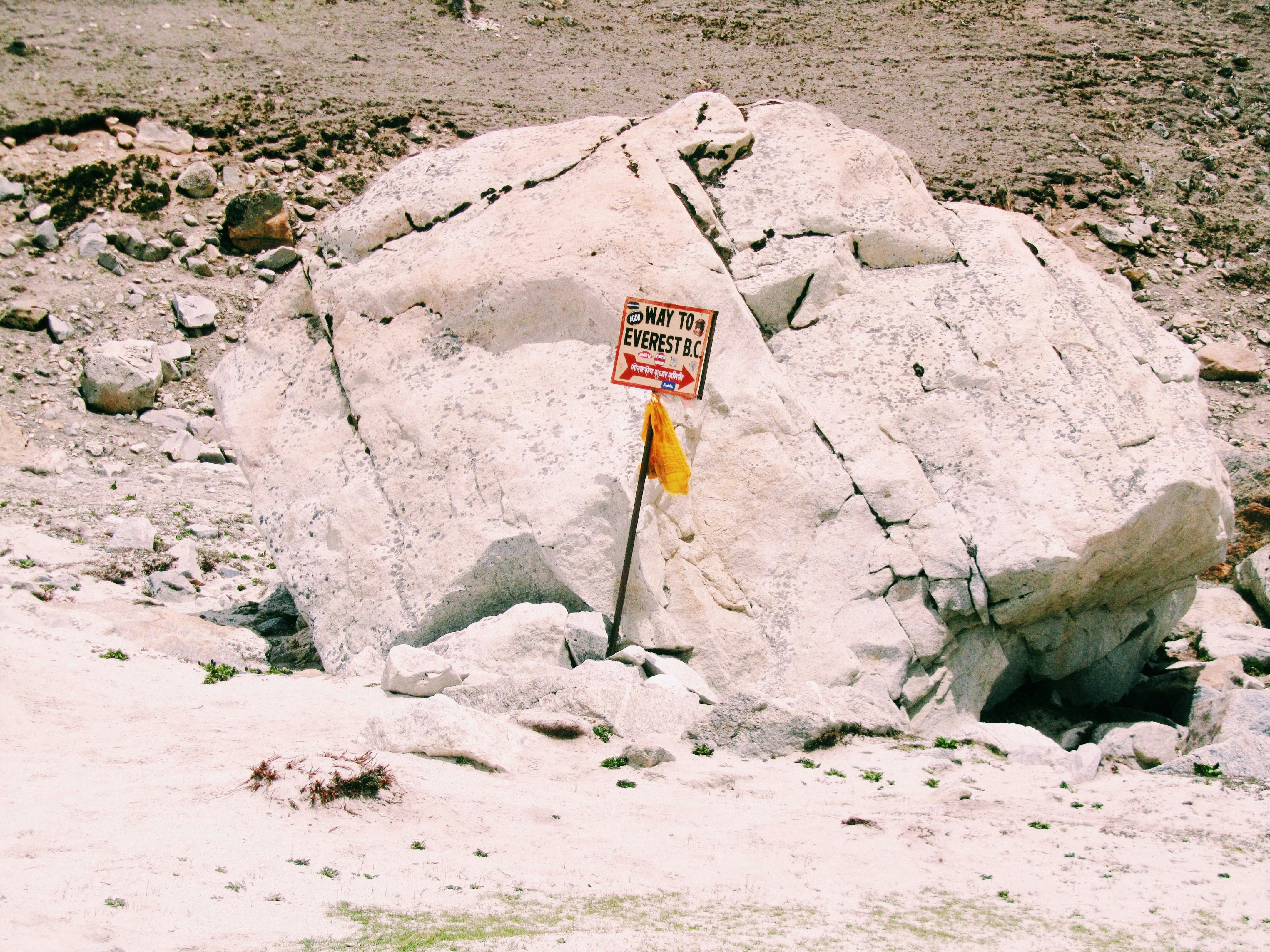 A weathered sign warns climbers about the dangers of Everest, positioned against a massive boulder in a rugged landscape.