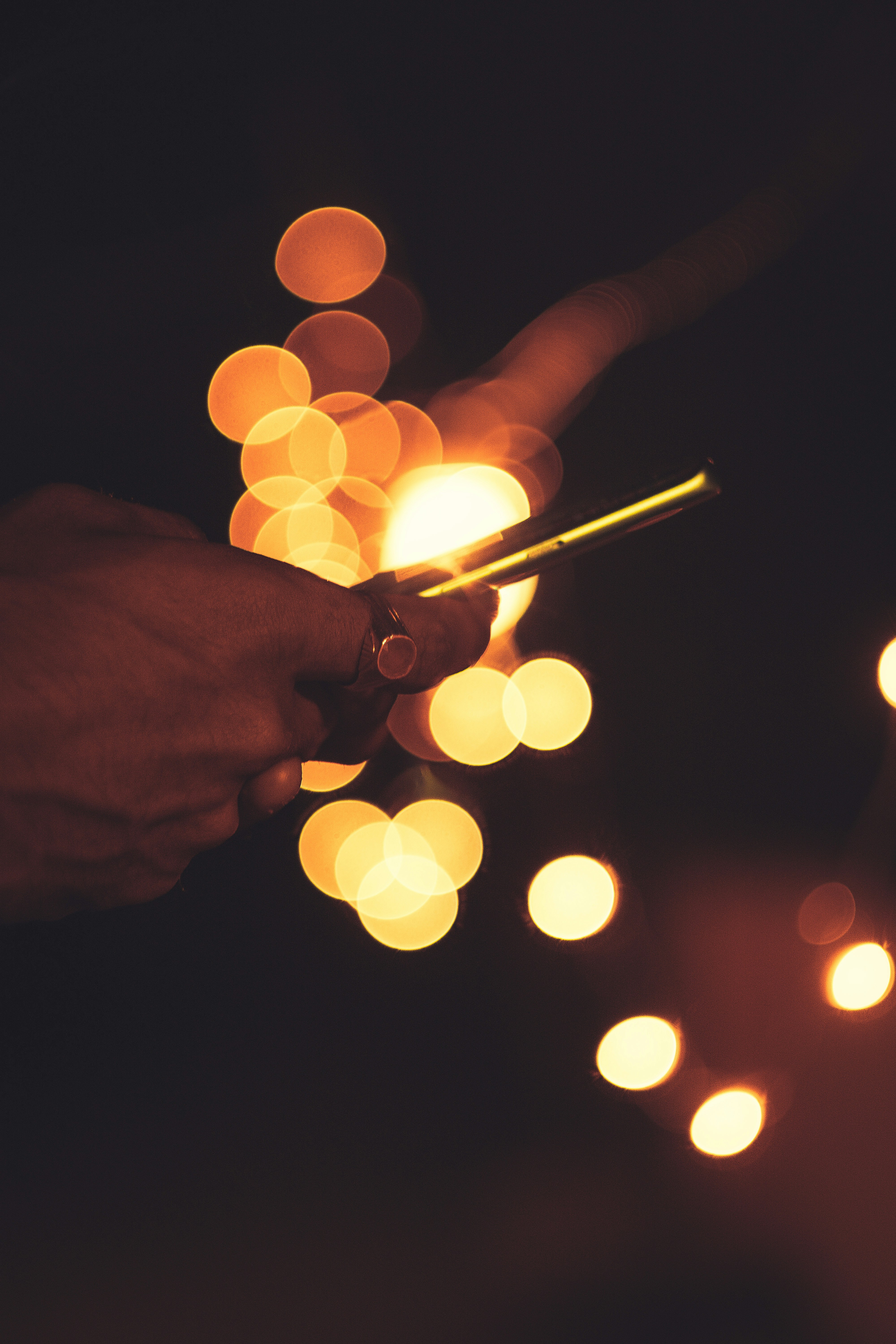 Close-up of a hand holding a sparkler, surrounded by a blur of glowing bokeh lights in a dark setting.
