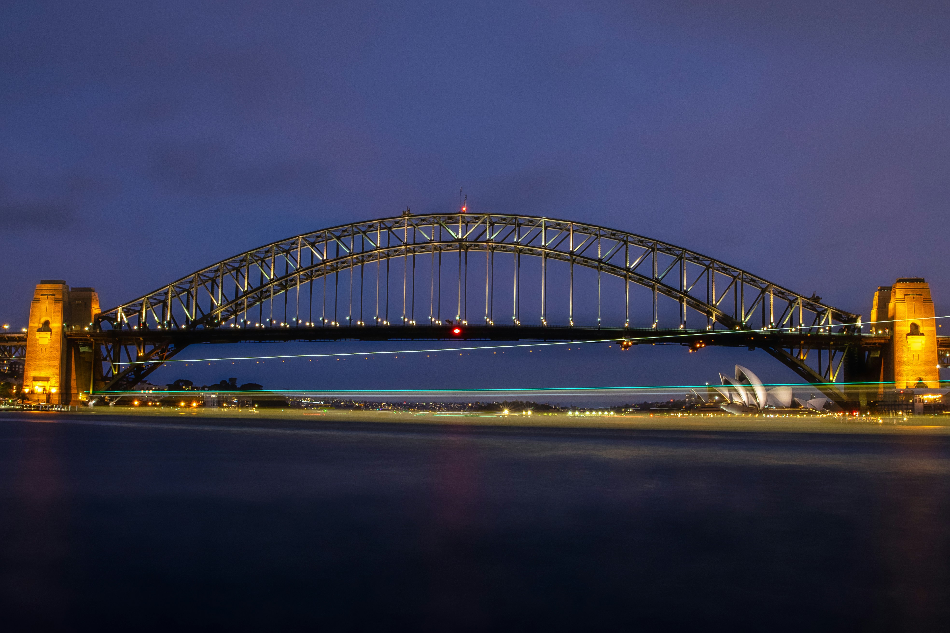 Sydney Harbour Bridge illuminated against a twilight sky, with the Sydney Opera House visible in the background. The scene captures the vibrant energy of the city at night.