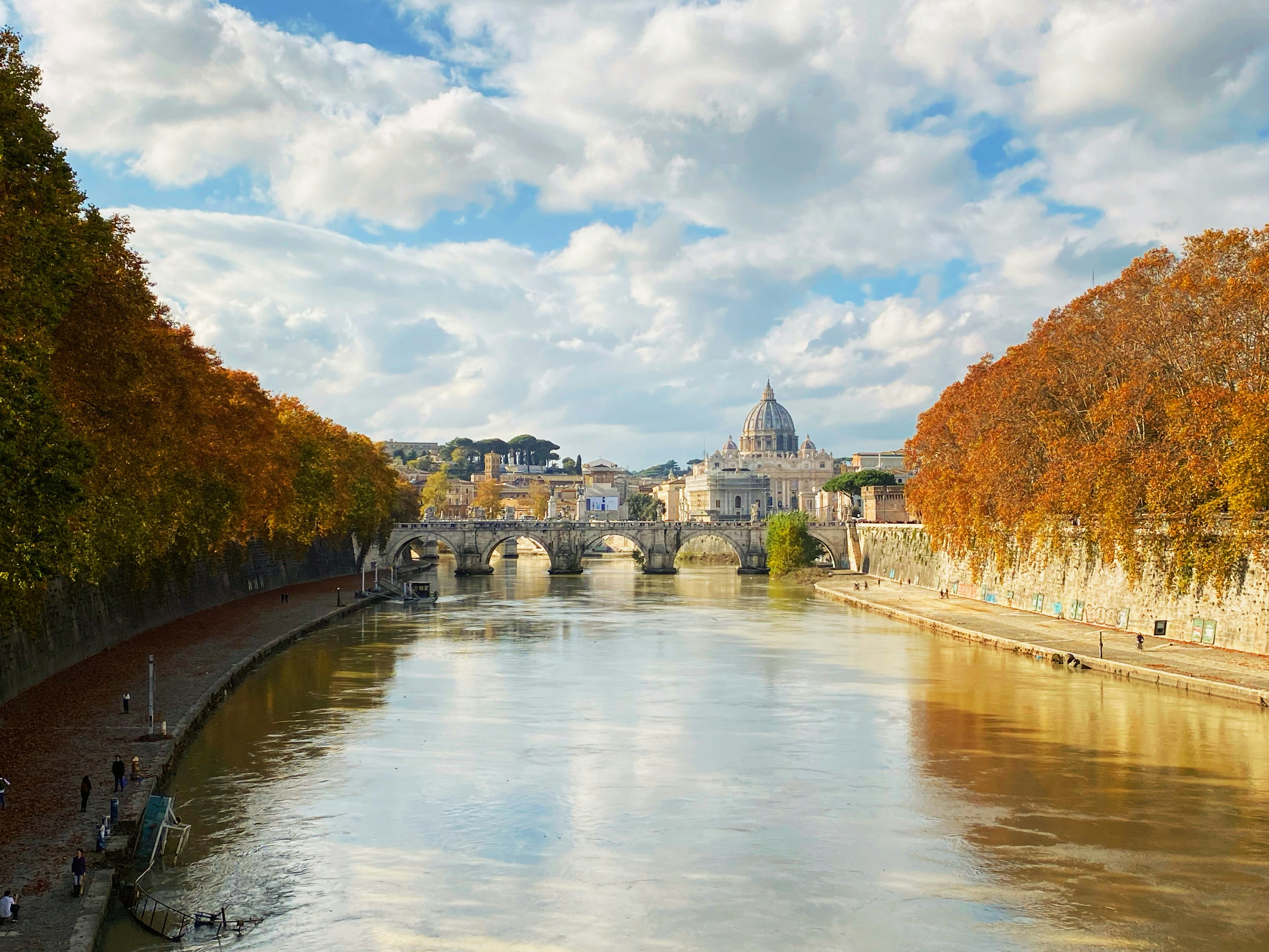 St. Peter's Basilica framed by vibrant autumn foliage along the Tiber River under a bright, cloud-dotted sky.