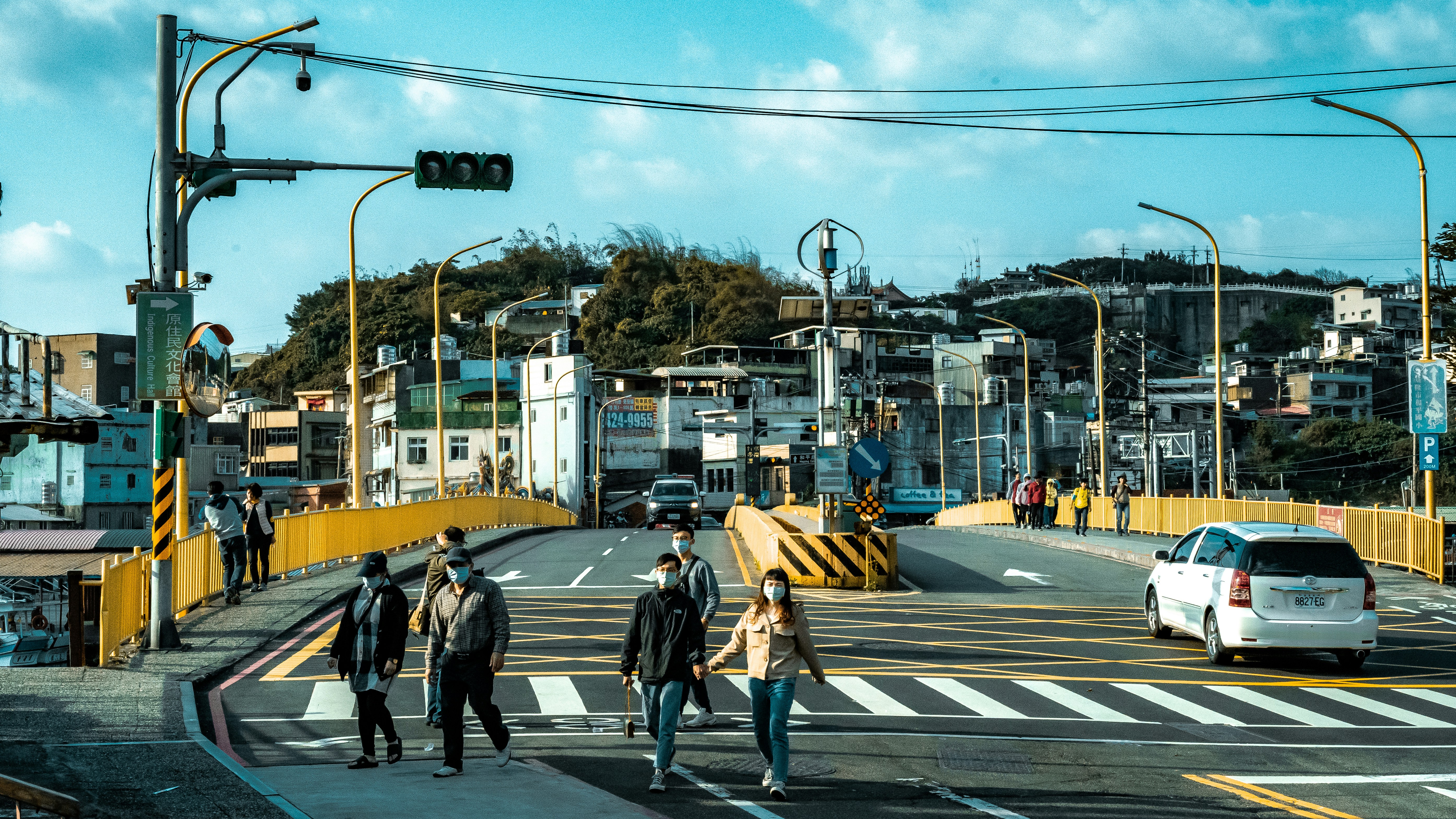 Pedestrians crossing a sunlit street with urban buildings in the background.