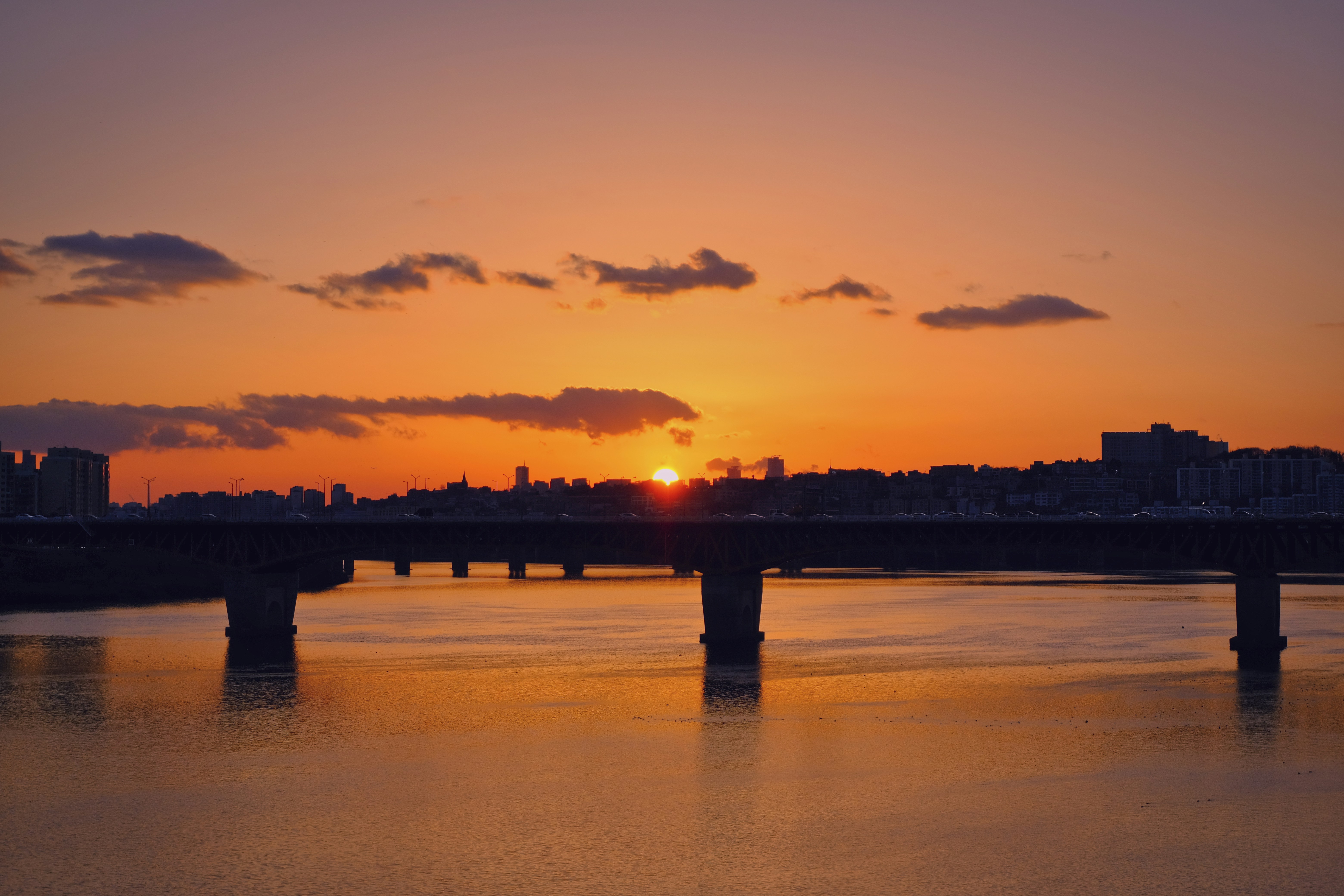 Golden sun setting behind a bridge, casting vibrant reflections on the water with a city skyline in the background.