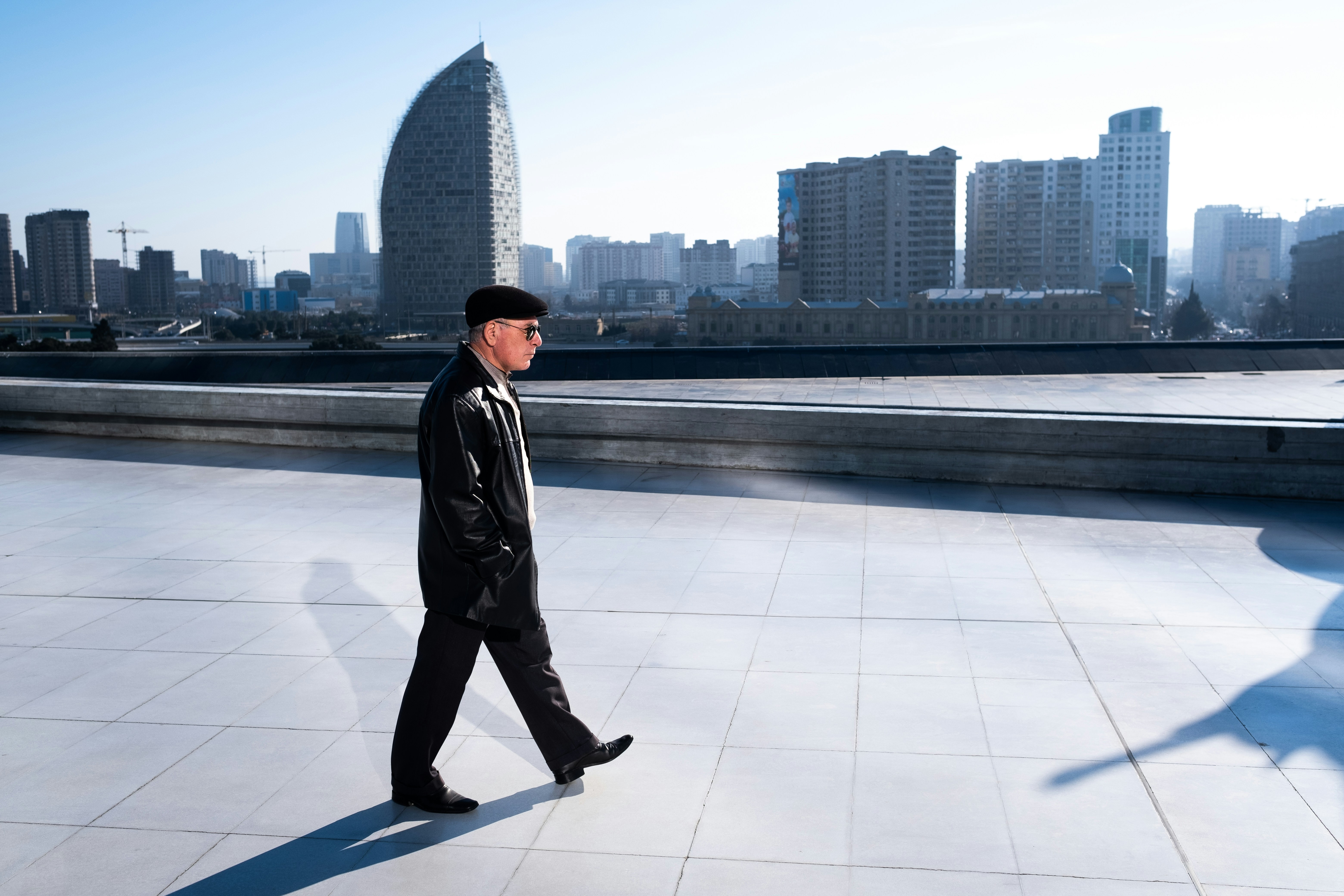 Man walking on a wide terrace with a city skyline and iconic curved building in the background.
