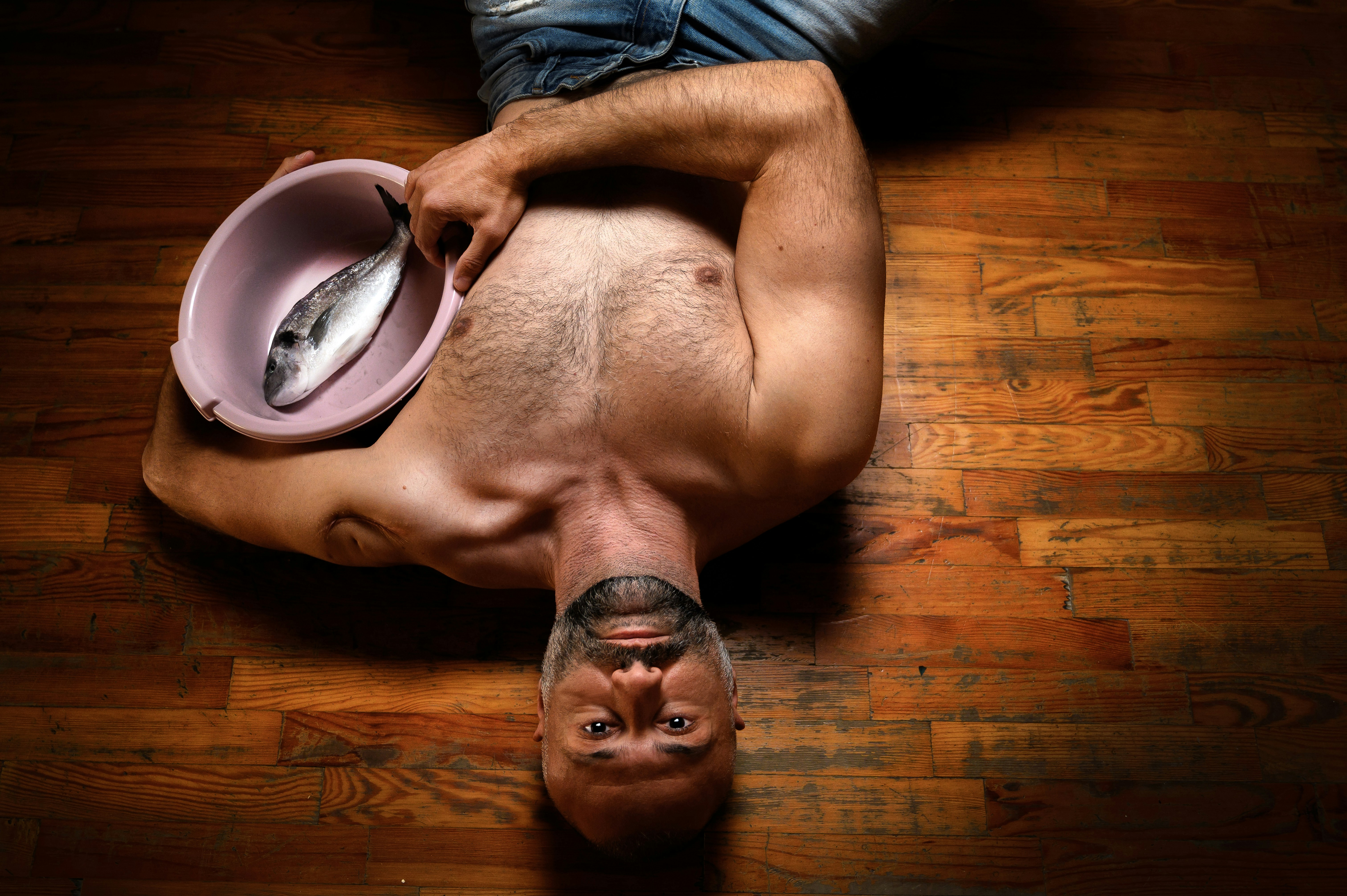 Topless man lying on a wooden floor holding a bowl.