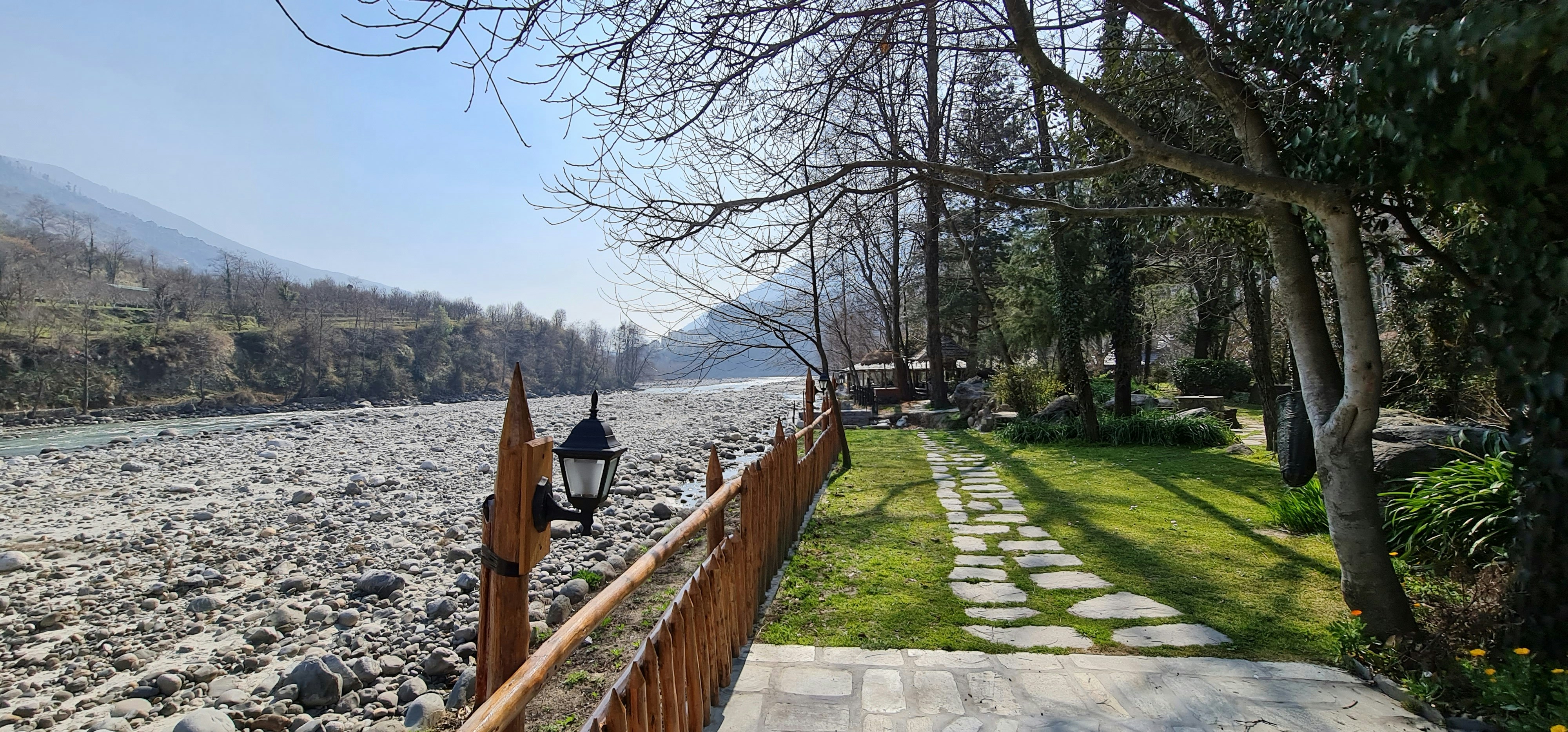 Sunlit path along a tranquil river with bare trees and lush grass.