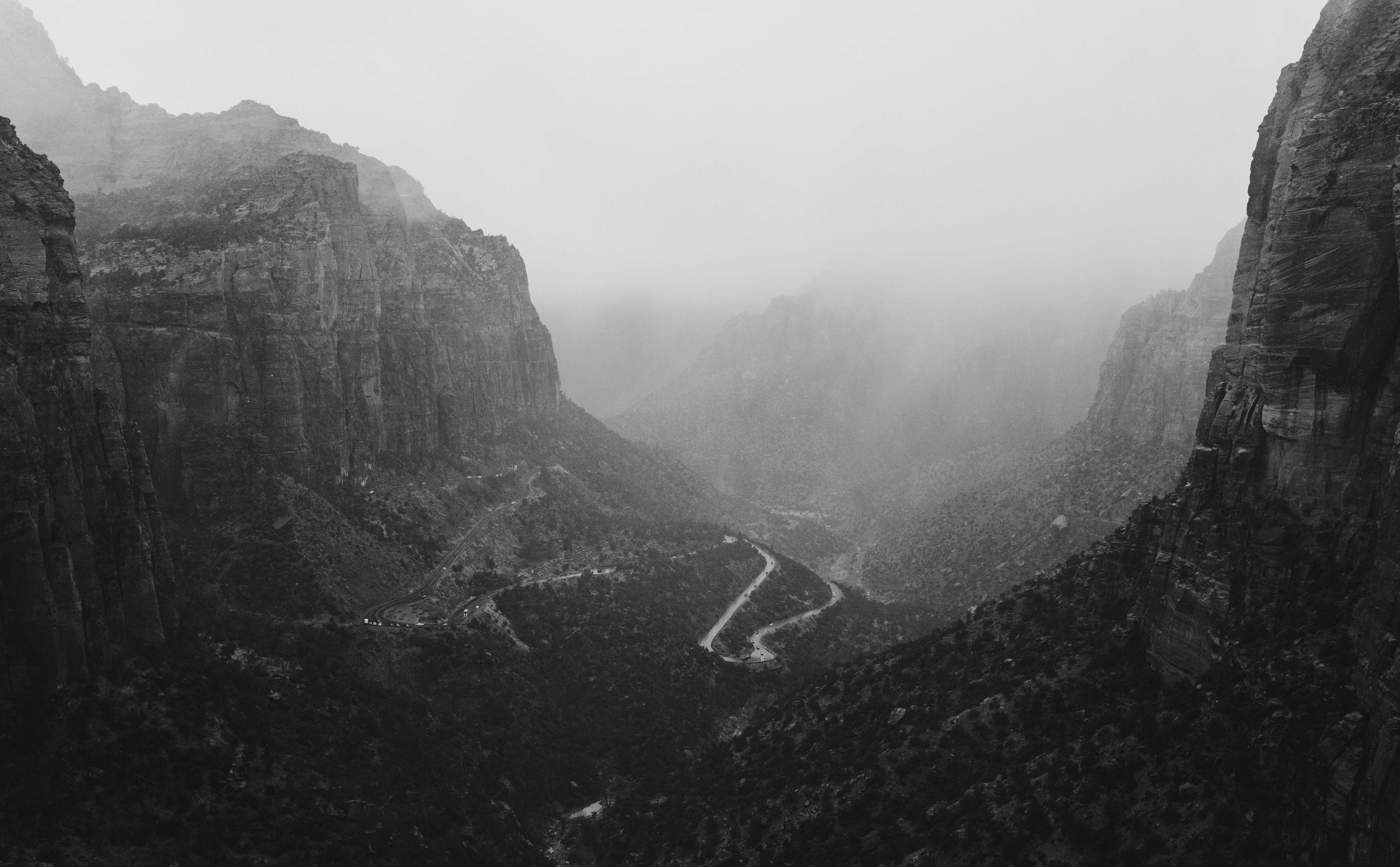 Grayscale view of towering cliffs enveloped in mist with a winding road below.