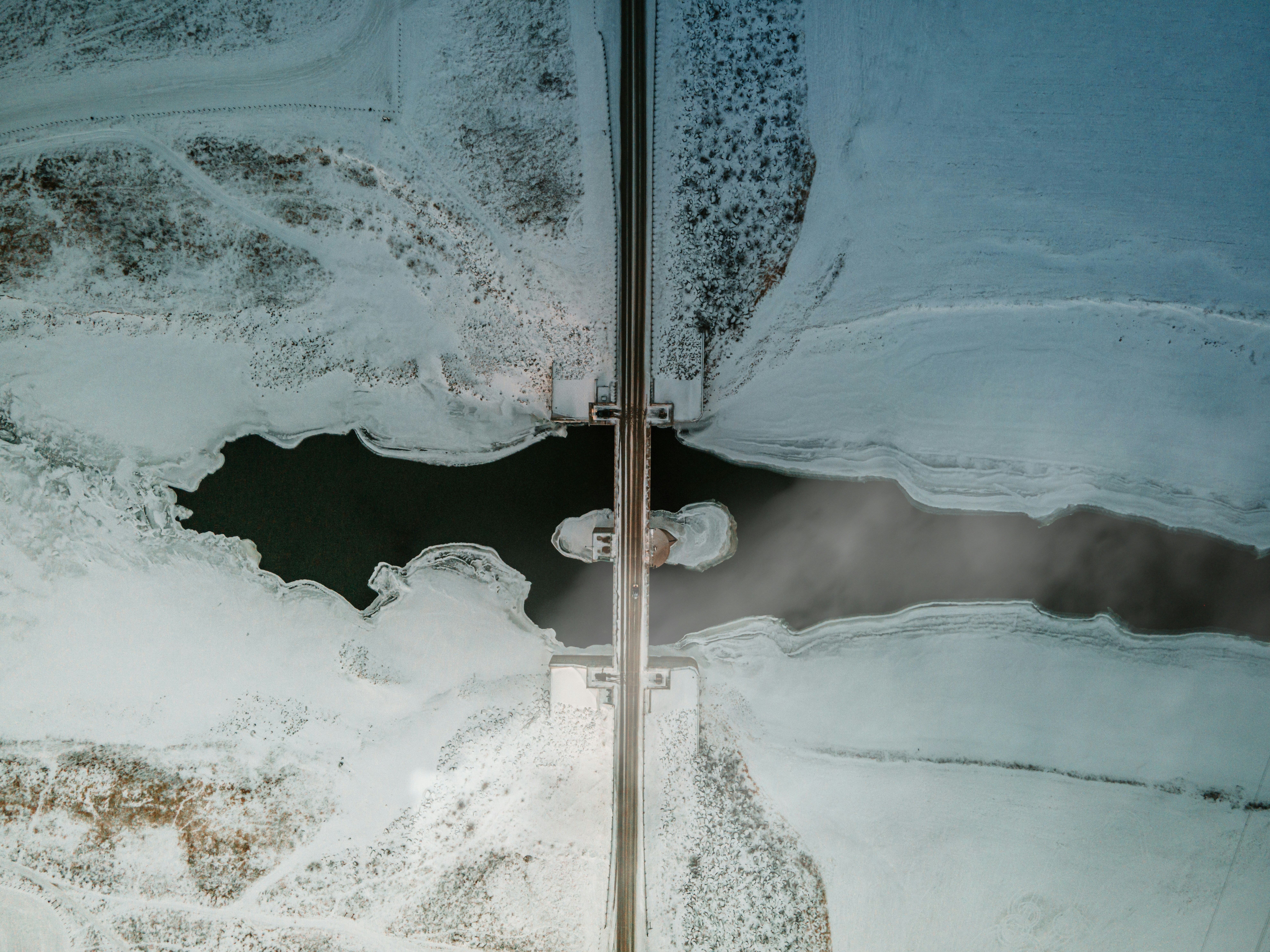 Aerial shot of a snow-covered landscape with a dark river cutting through, flanked by a straight road.