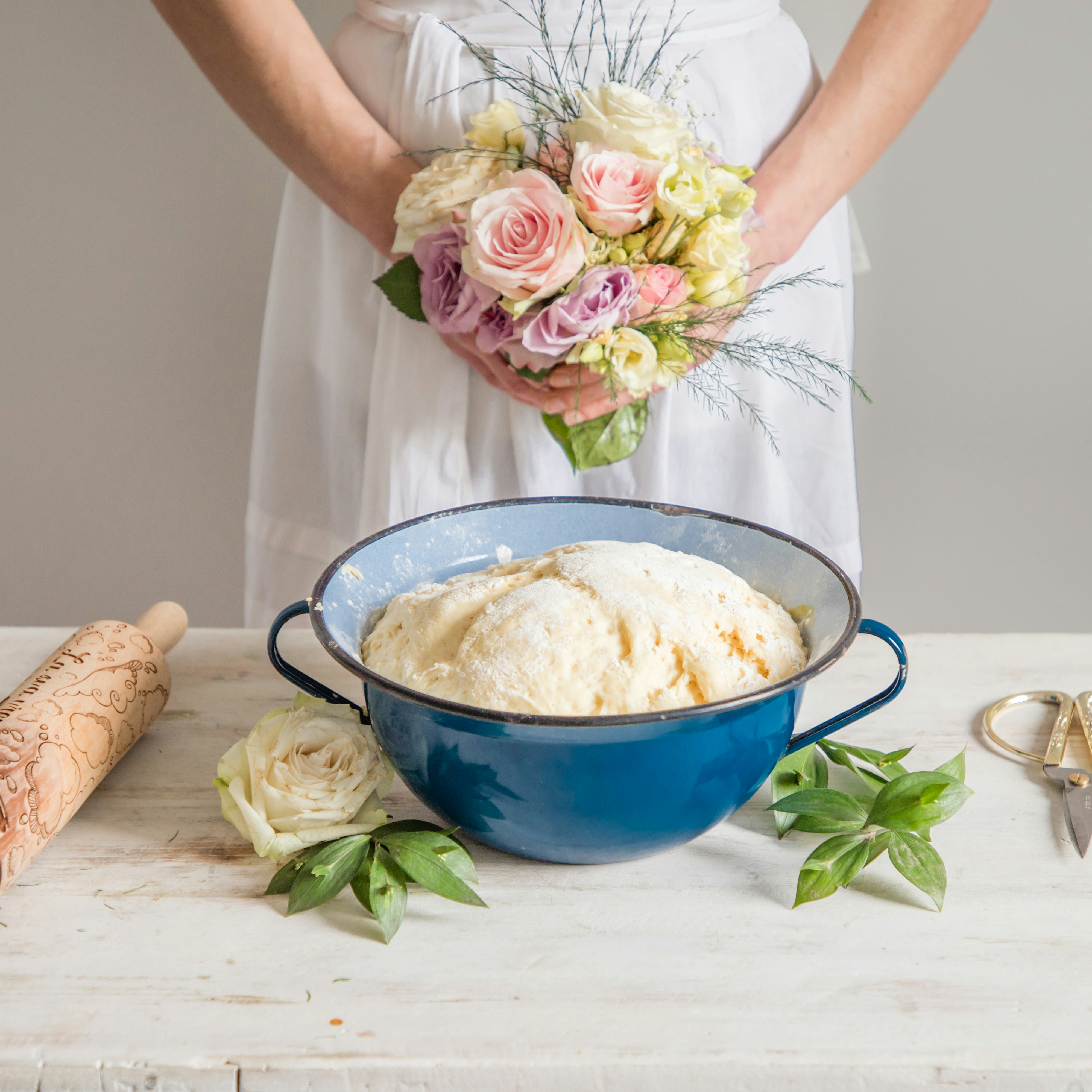 A person in a white apron holds a bouquet of pastel roses while standing over a blue bowl filled with dough, accompanied by a rolling pin and fresh flowers on a wooden table.