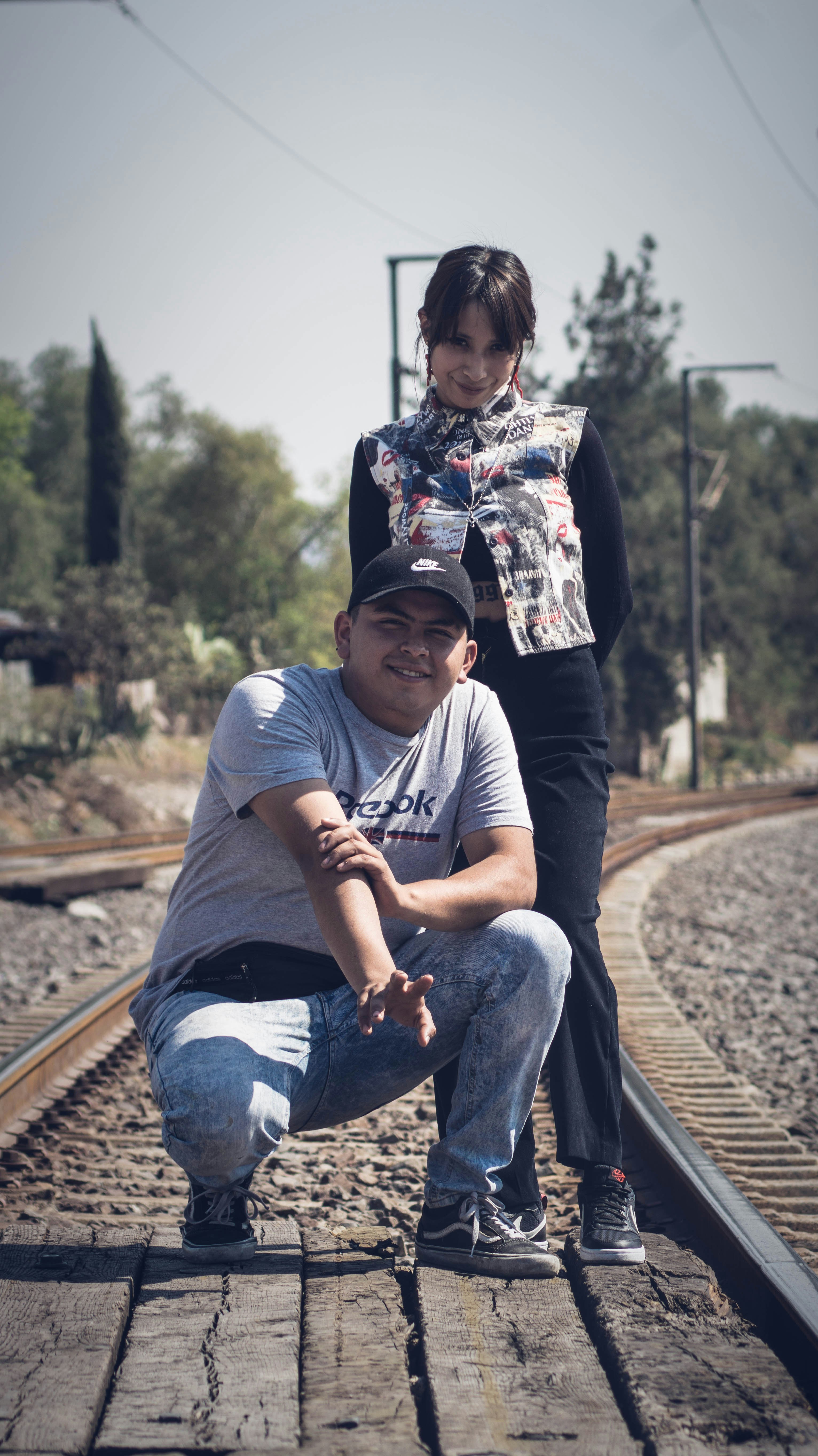 Two friends posing on a wooden railway track, exuding a casual and confident vibe against a backdrop of greenery.