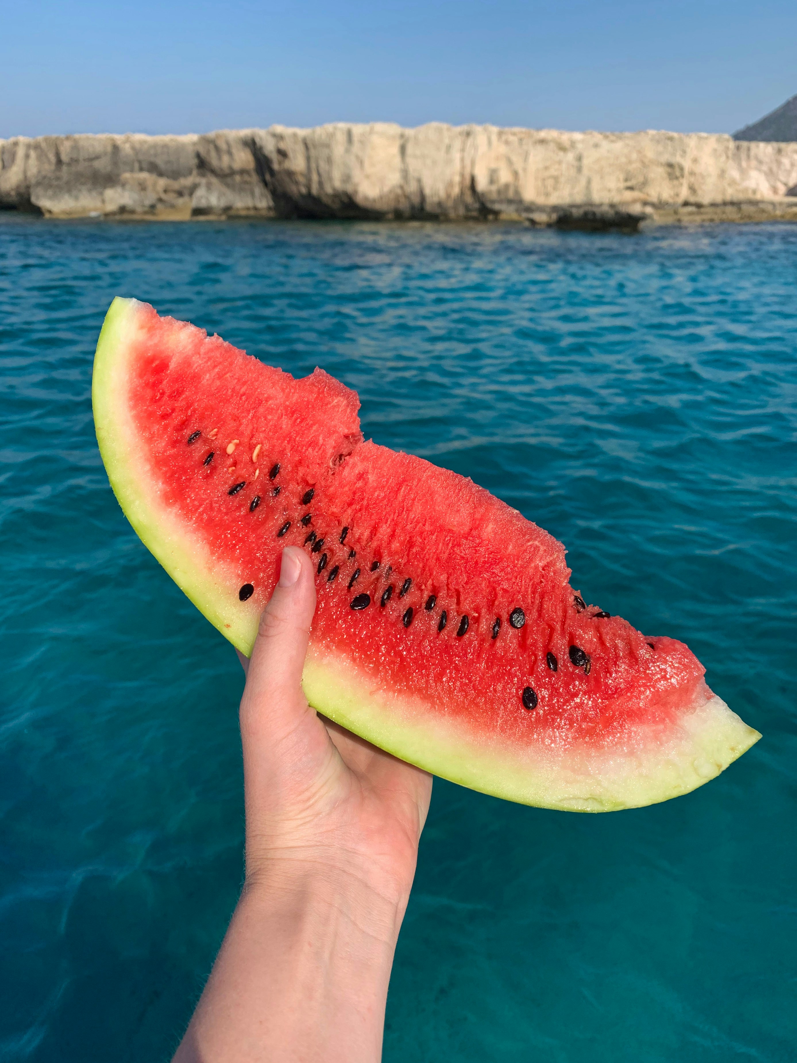 Hand holding a vibrant slice of watermelon against a backdrop of clear blue water and rocky coastline.