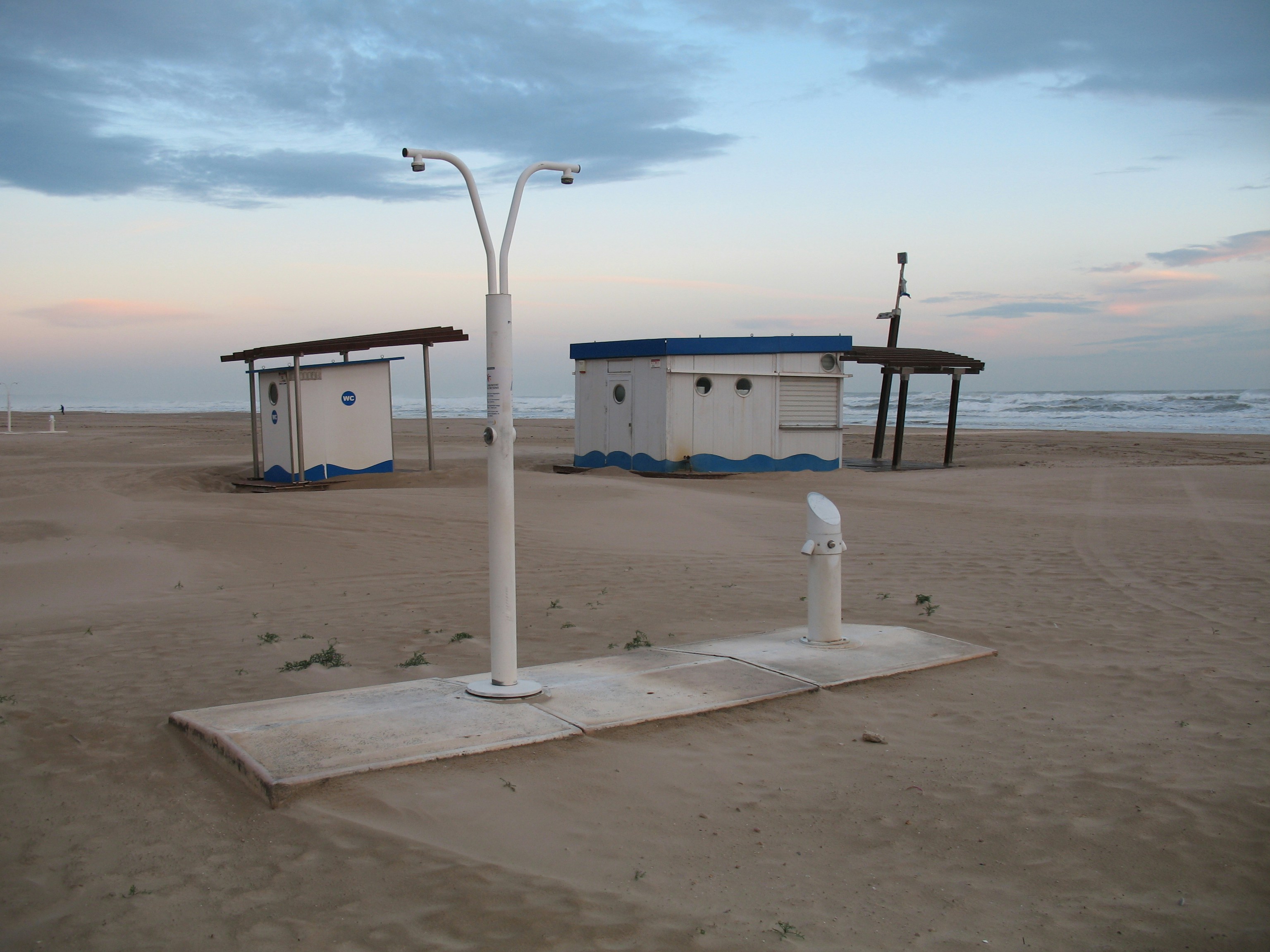 Beach showers and changing huts stand on a sandy shore as the sun sets, creating a serene atmosphere. The scene captures the essence of coastal relaxation.